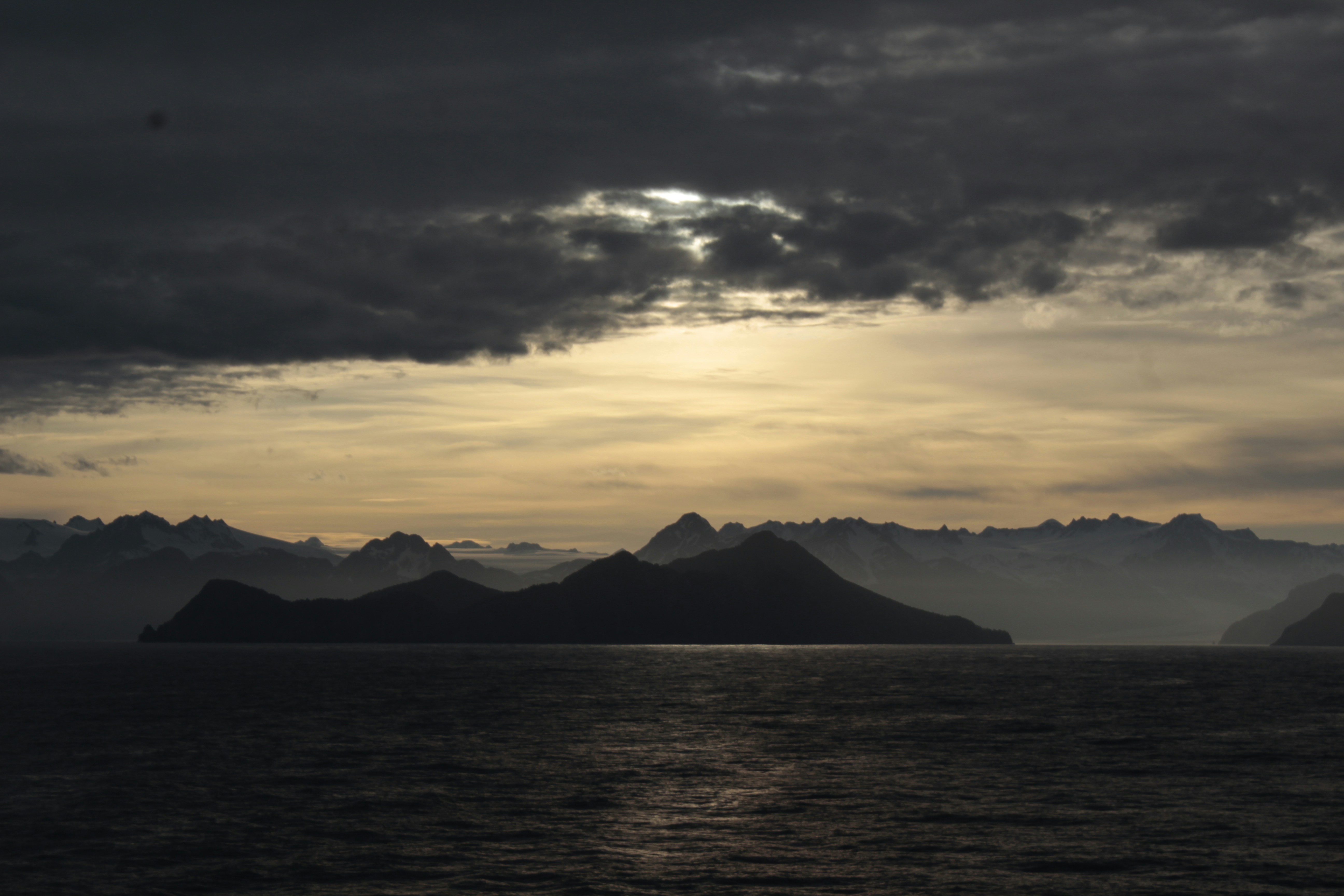 Silhouetted mountains rise against a moody sky, reflecting muted light on tranquil waters. The scene evokes a sense of calm and introspection.
