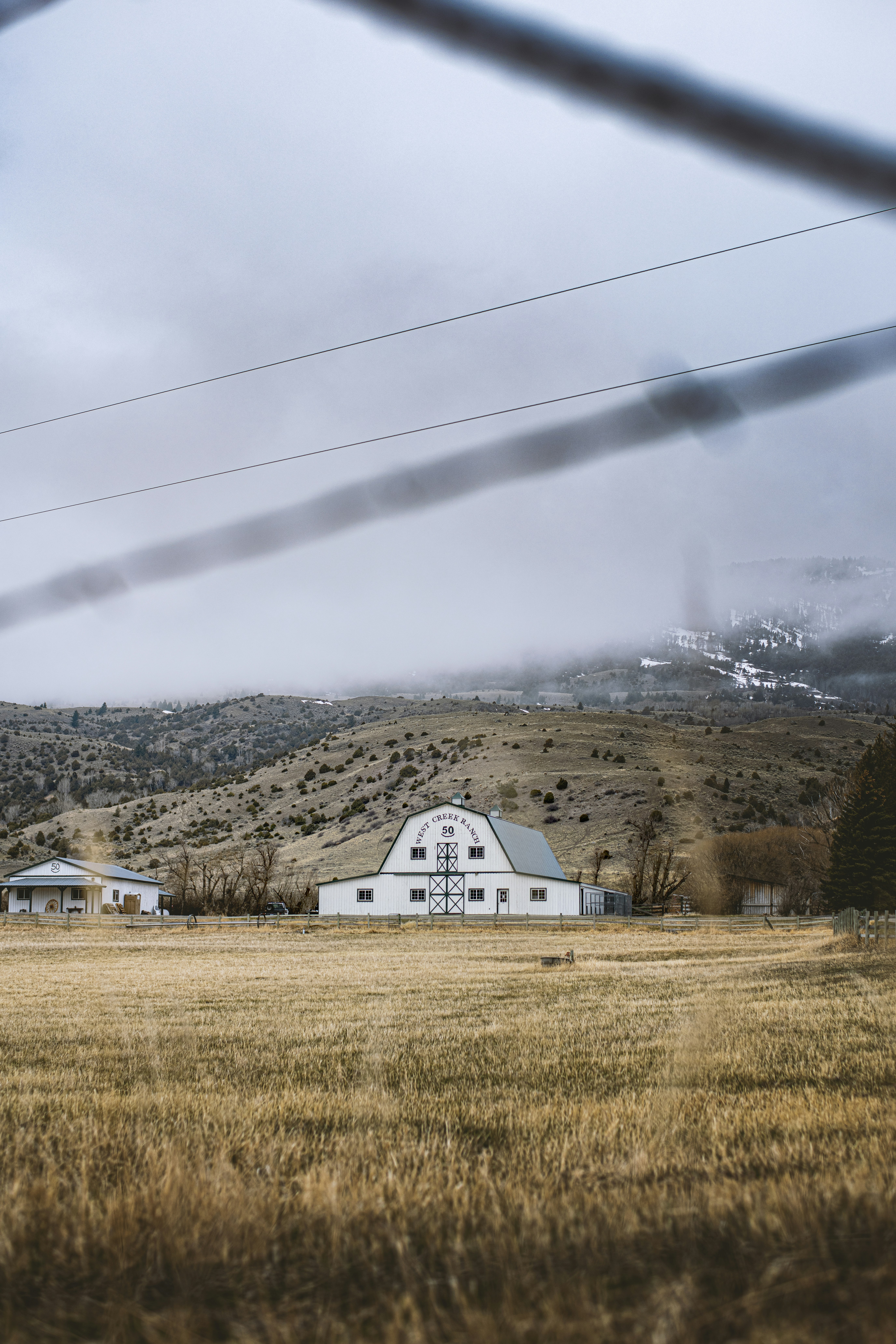 A vintage barn stands prominently against a backdrop of rolling hills and misty mountains, framed by a barbed wire fence. The scene evokes a sense of nostalgia and tranquility.