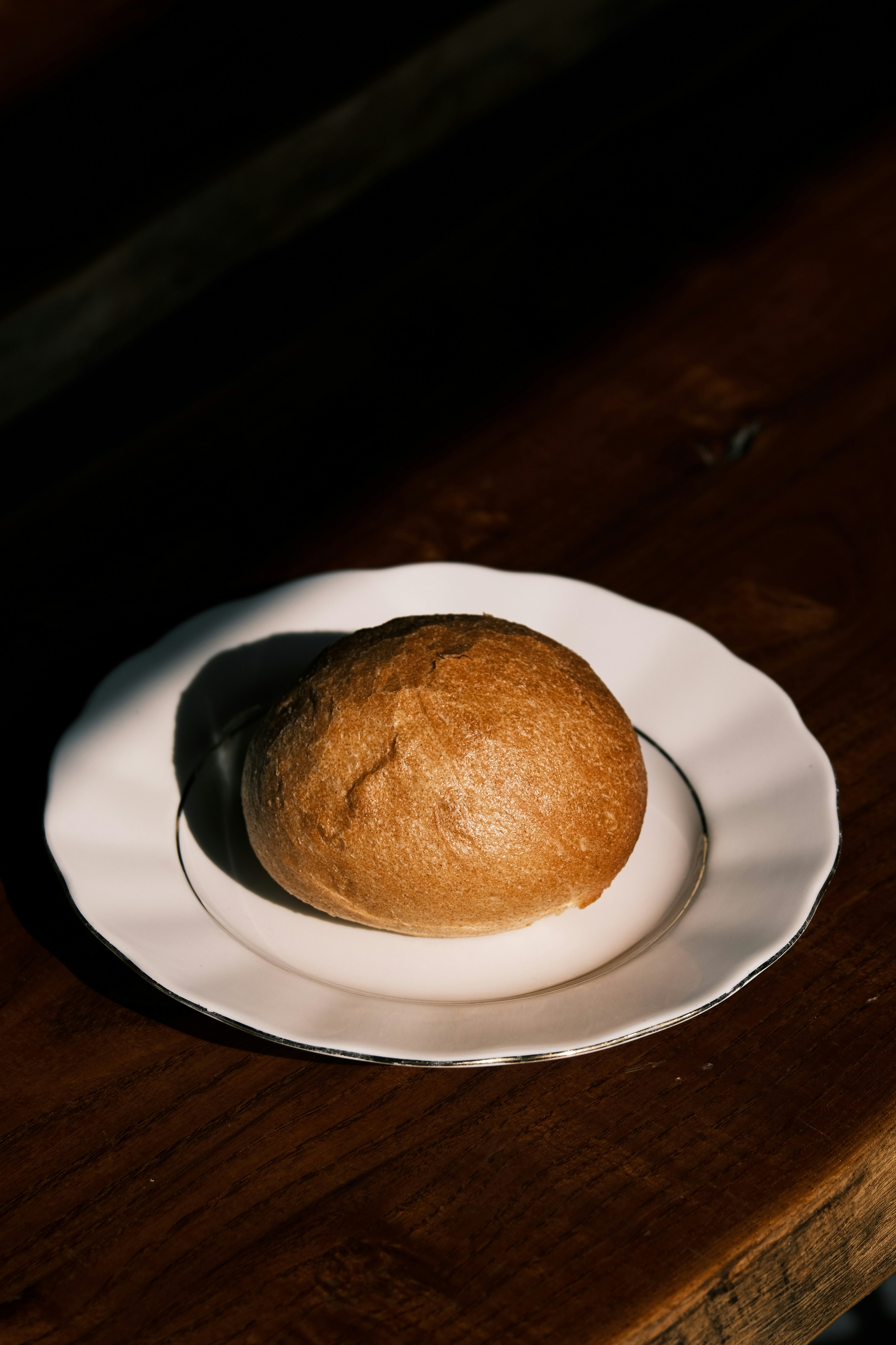A freshly baked bread roll resting on a delicate plate, illuminated by soft light against a wooden backdrop.
