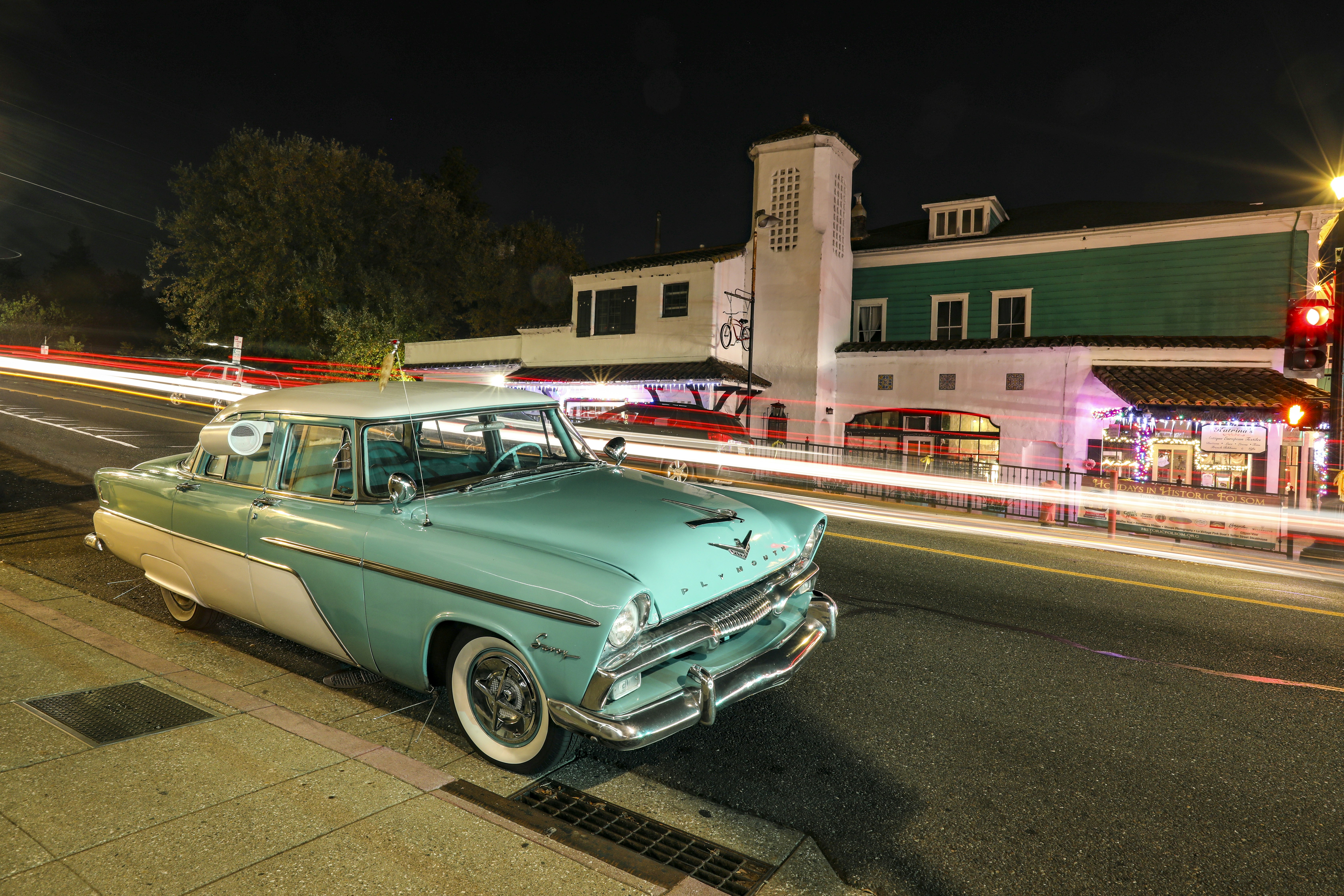 Classic turquoise car parked on a city street at night with light trails from passing vehicles.