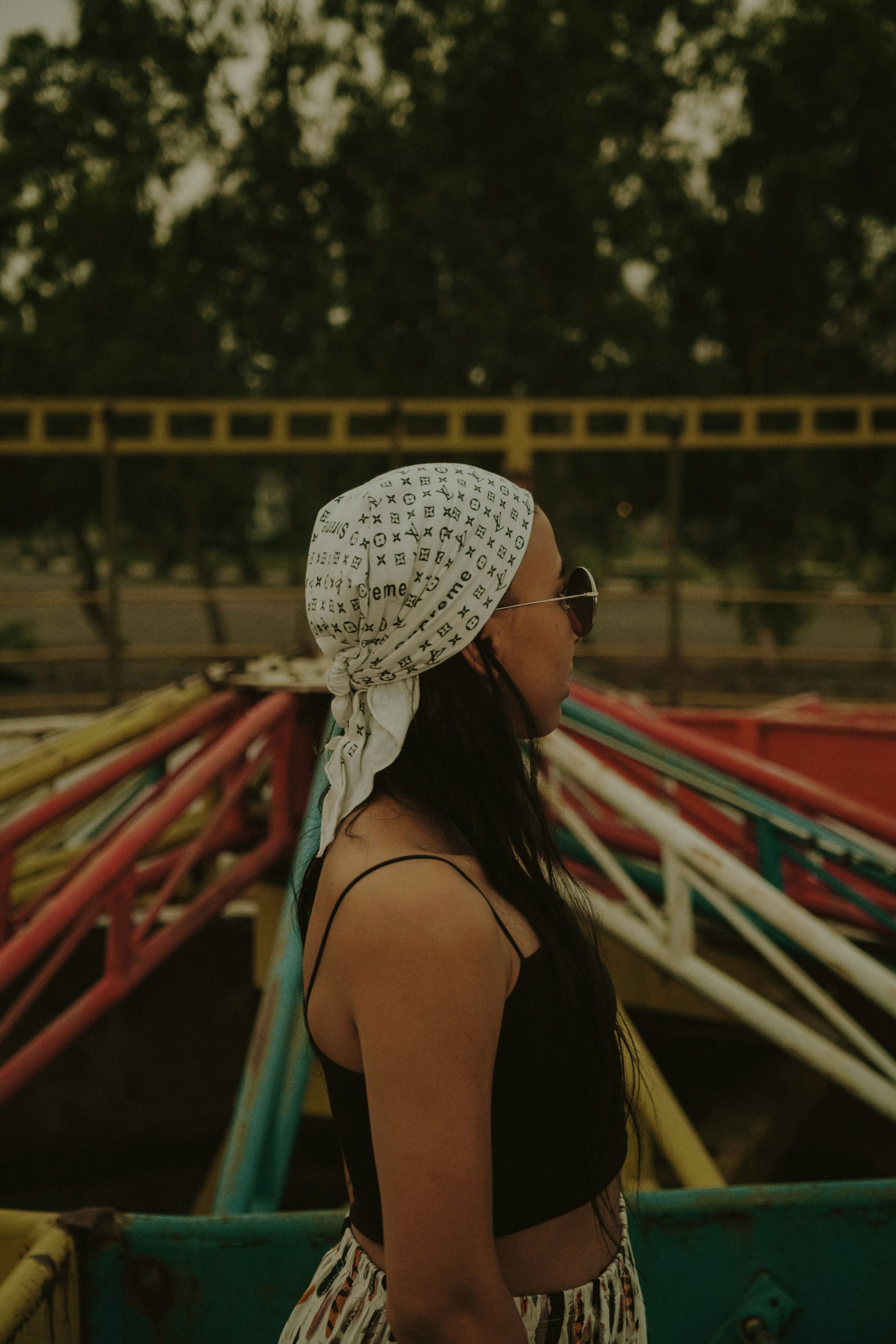 A woman in a patterned headscarf and sunglasses stands against a colorful backdrop of geometric shapes, embodying a blend of urban style and youthful spirit.