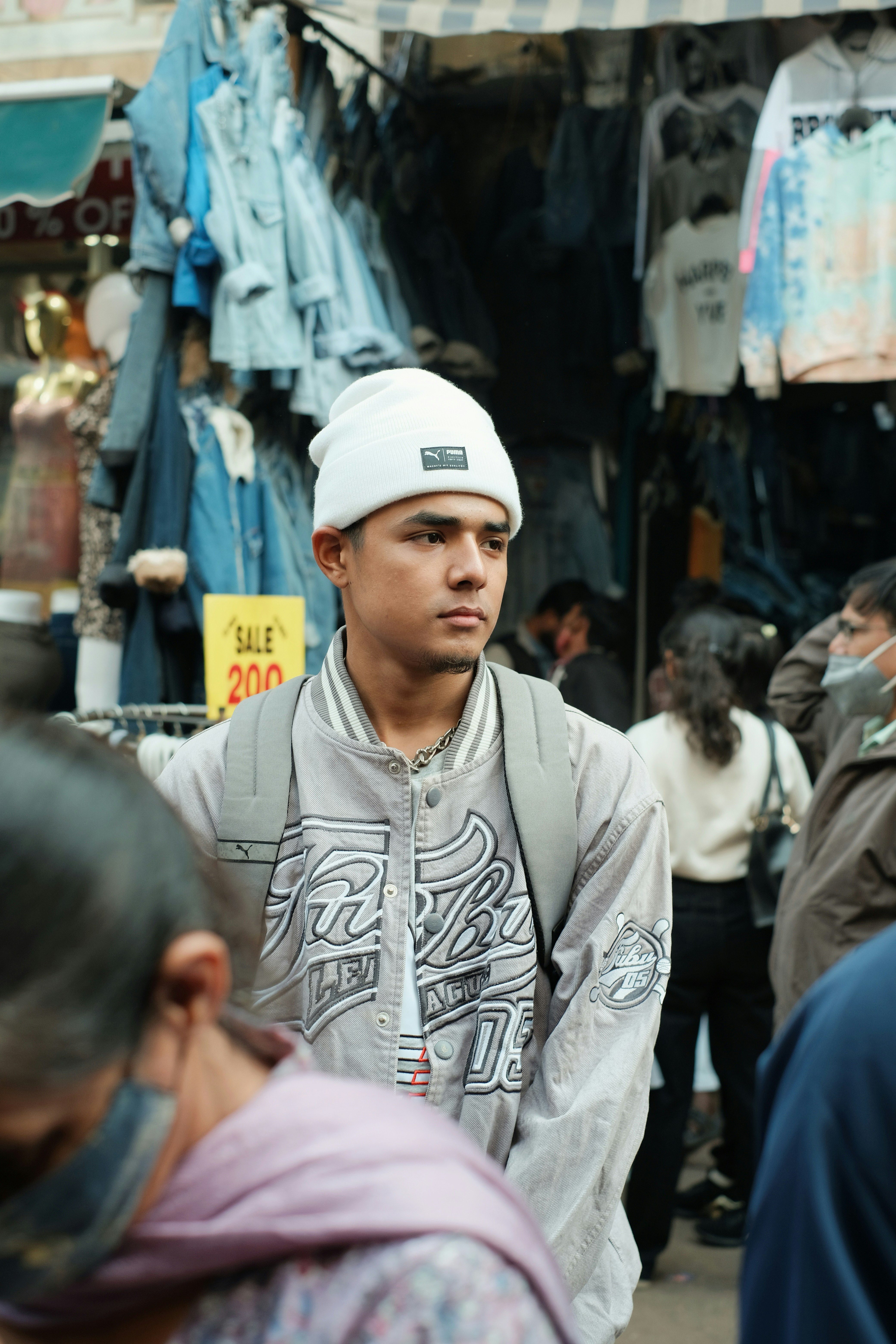 Young man in a light jacket and beanie stands amidst a bustling market, surrounded by hanging clothing and shoppers. The scene captures the essence of urban life.