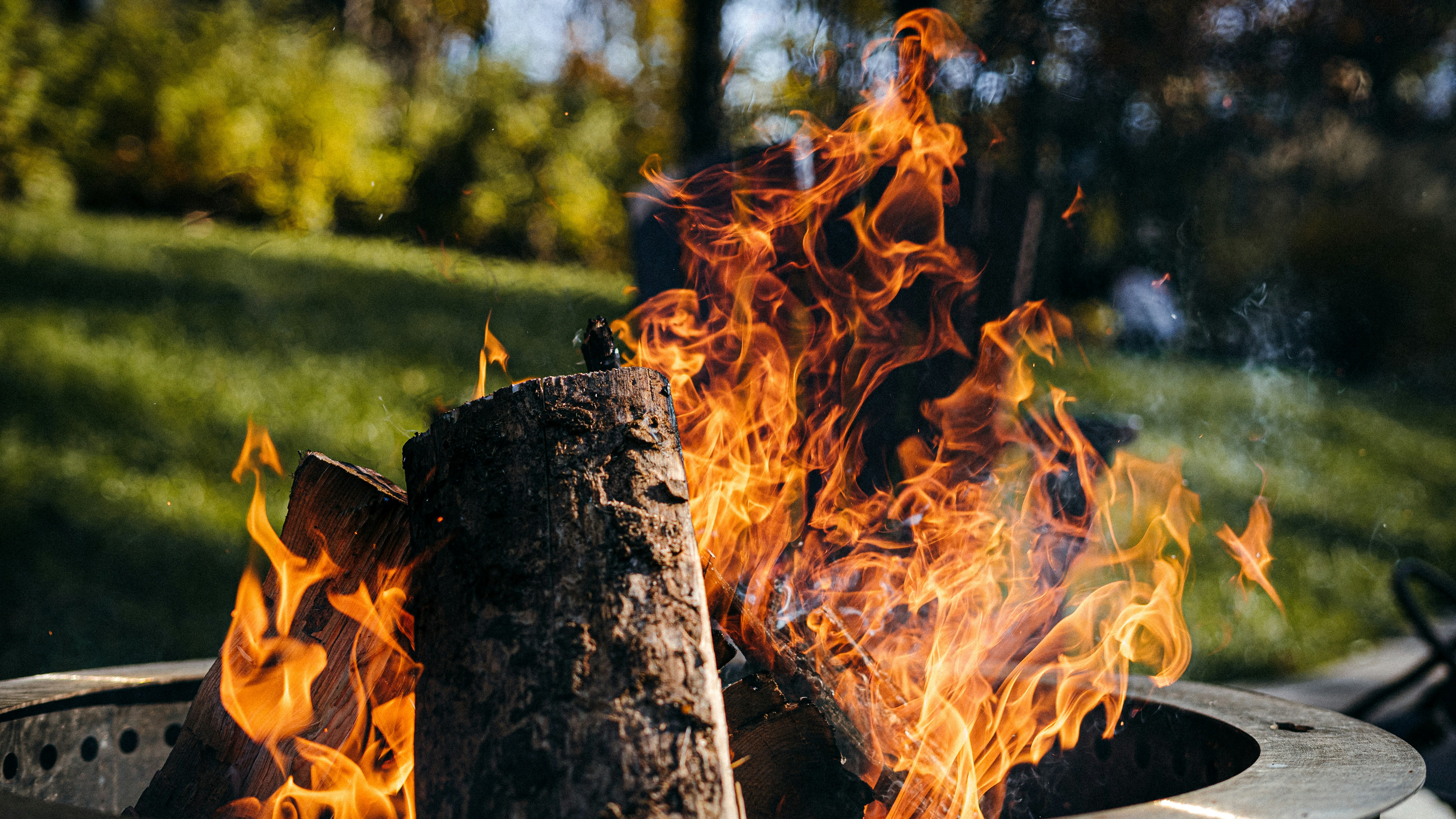 Flames flicker and swirl around logs in a fire pit, capturing the essence of a cozy outdoor gathering. The vibrant colors of the fire contrast with the lush green background.