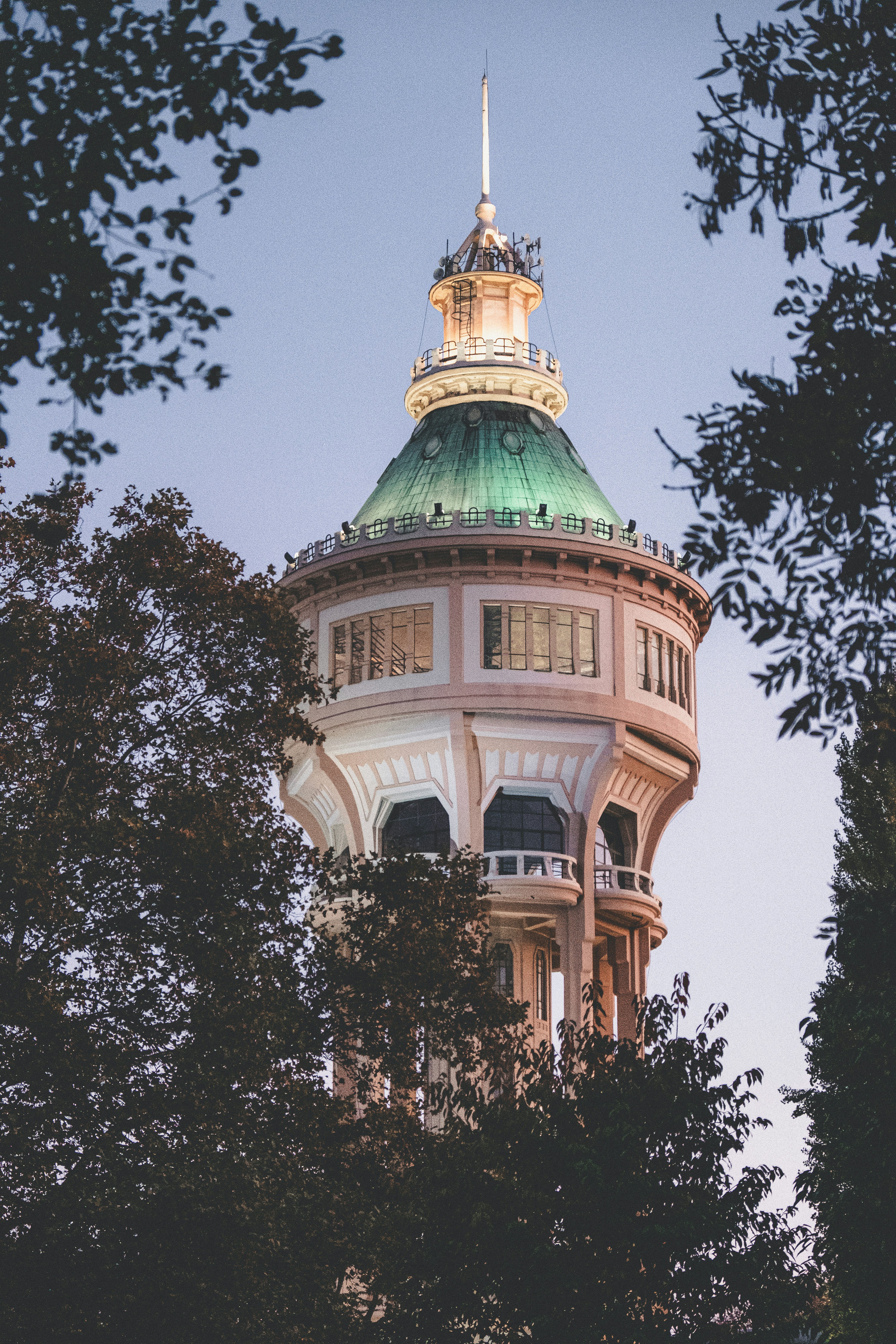 Historic tower adorned with a green dome, framed by lush trees during twilight. Illuminated windows enhance its architectural beauty.