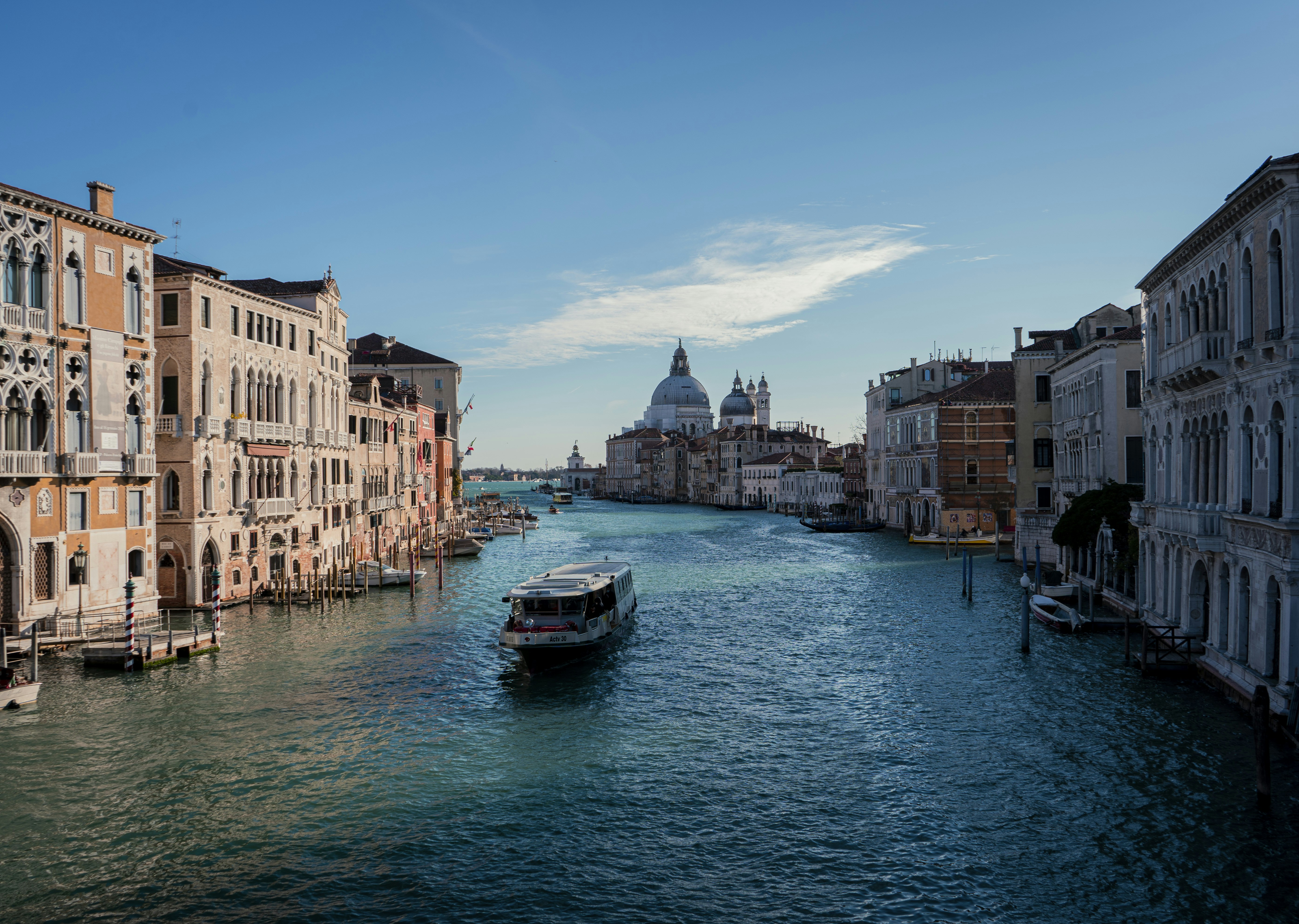 A scenic view of a serene canal in Venice, showcasing historic buildings lining the waterway with a boat gliding through. A clear blue sky enhances the tranquil atmosphere.