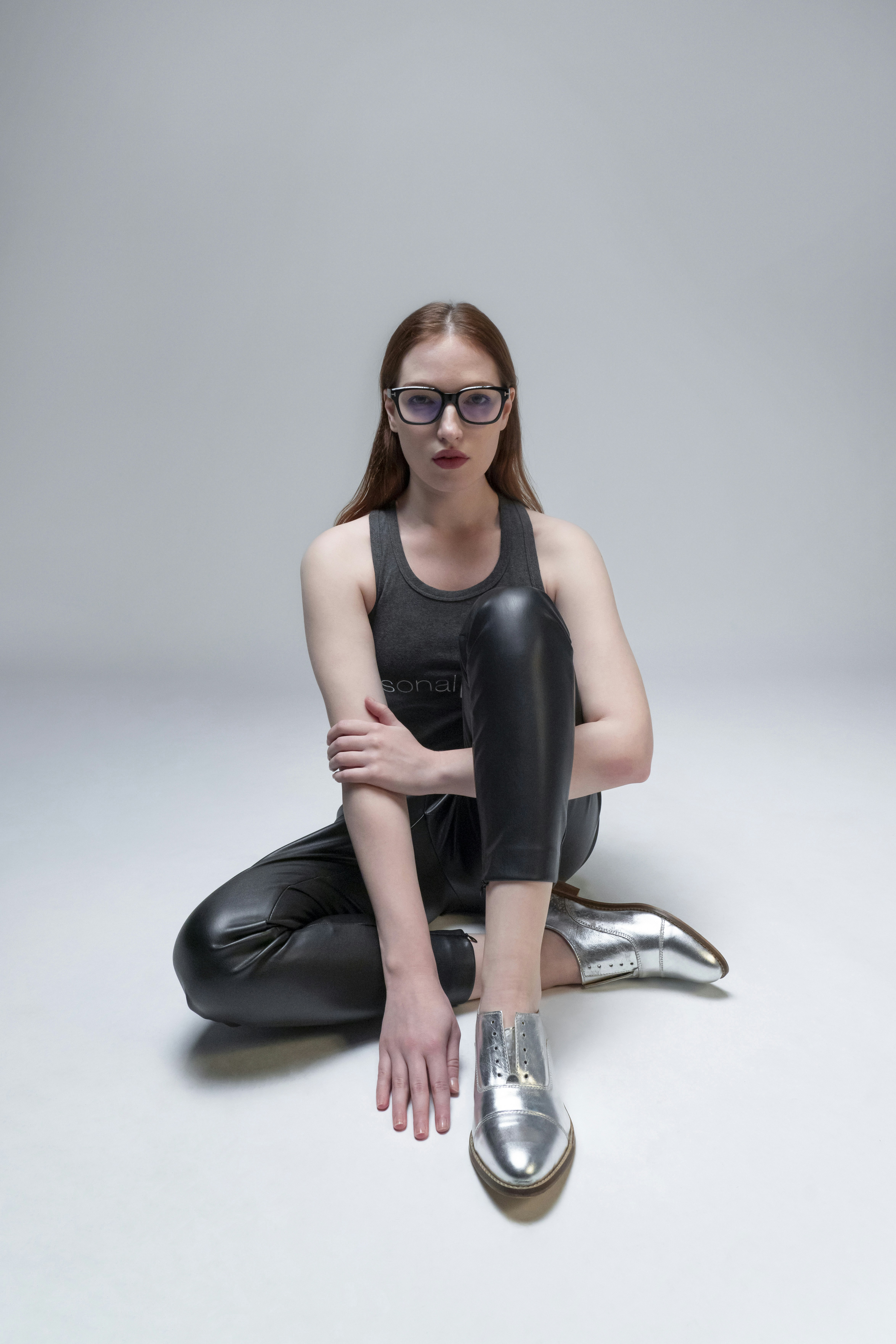 Model seated on the floor, showcasing a stylish outfit with silver shoes and black leather pants against a neutral backdrop.