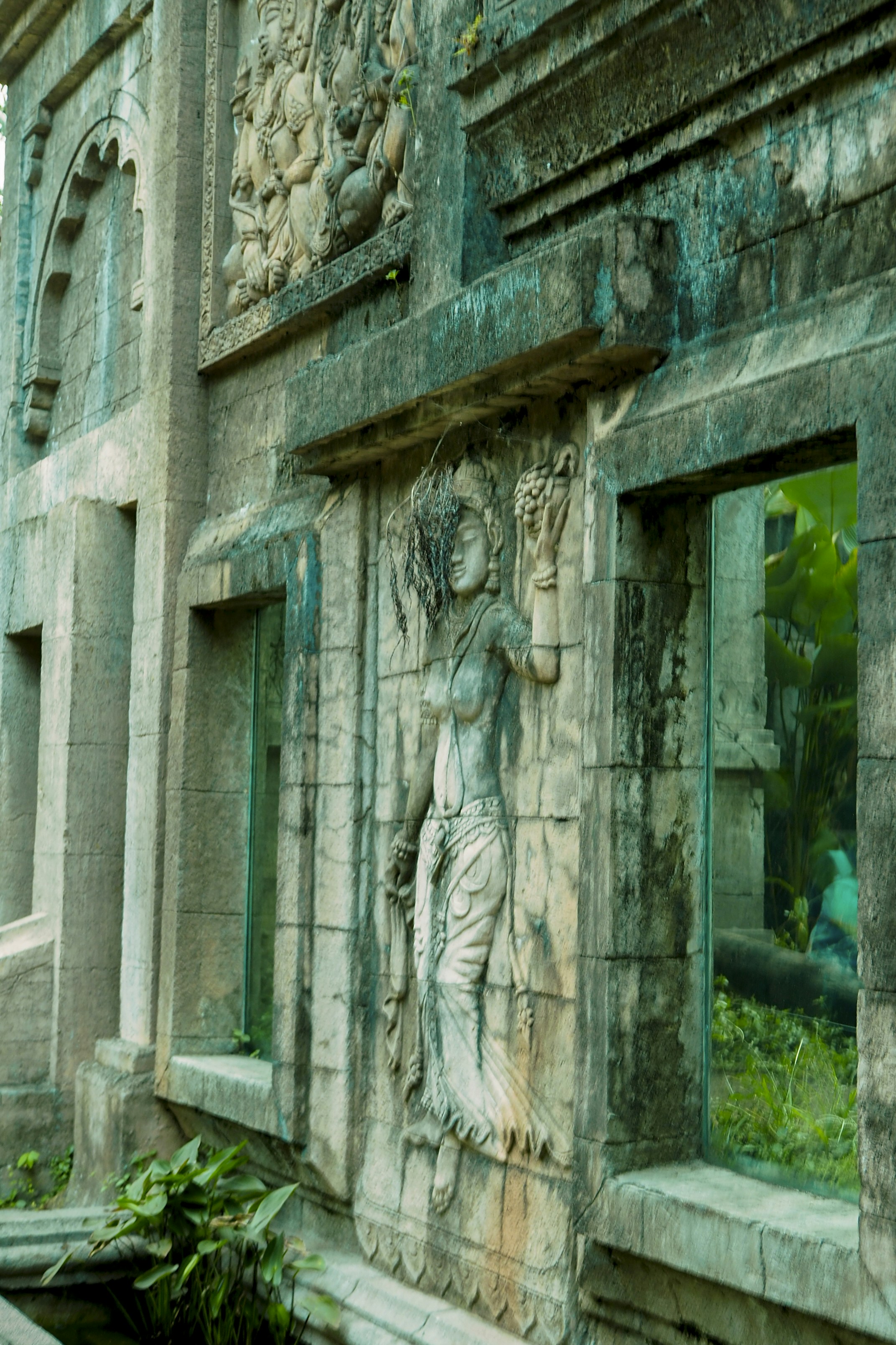 Intricate stone carving of a figure holding fruits, framed by a weathered wall with lush greenery visible through glass windows.