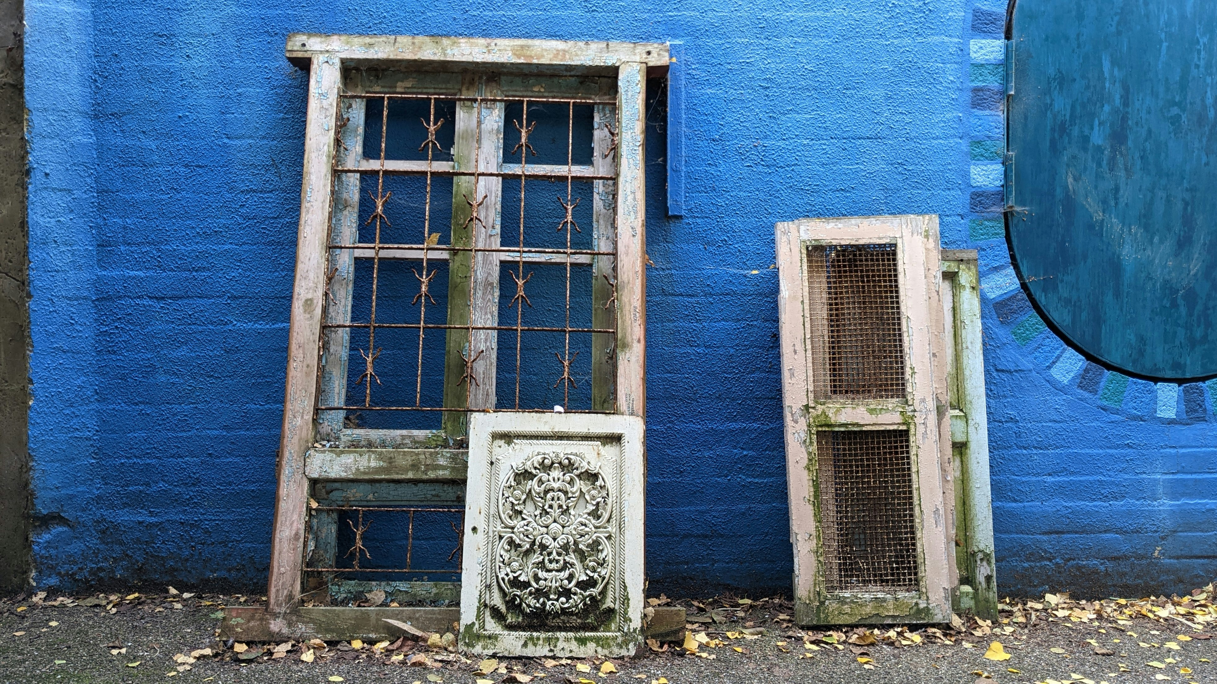 Weathered wooden doors and a decorative panel lean against a vibrant blue wall, showcasing a blend of textures and colors.