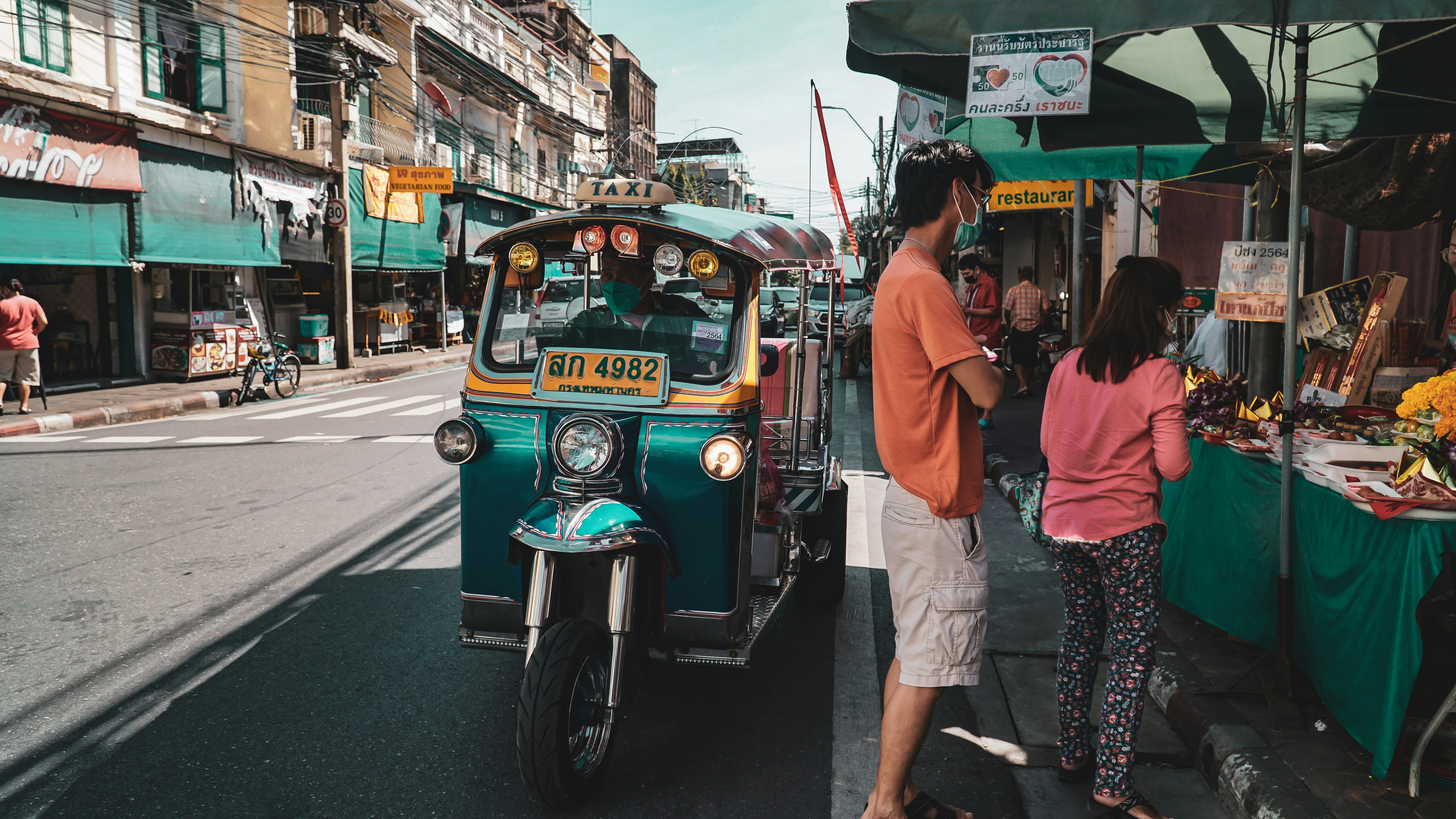 Colorful tuk-tuk parked beside a bustling Bangkok street market, with people exploring fresh produce stalls under a bright sky.