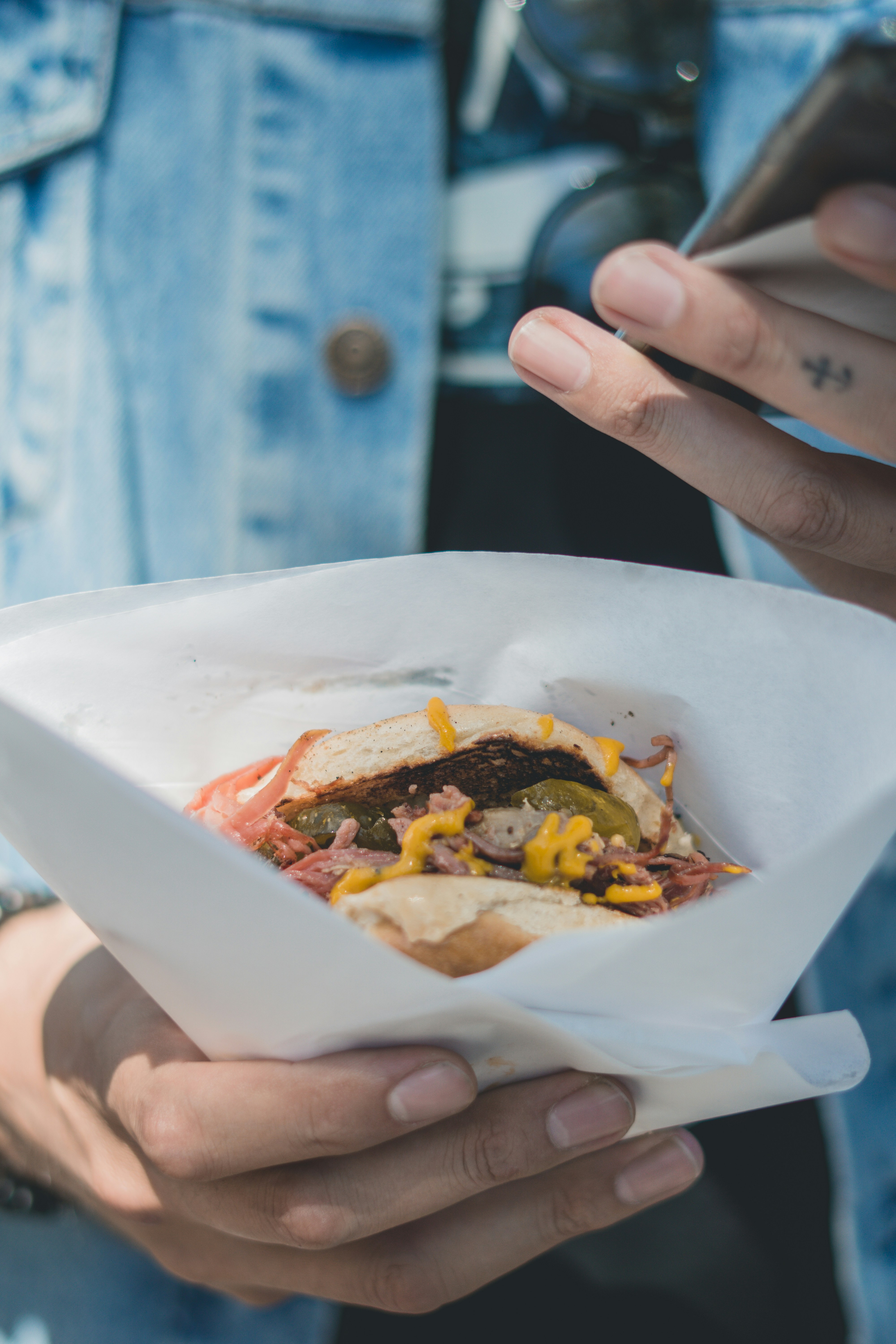 Person holding a wrapped sandwich with toppings in a casual outdoor setting.
