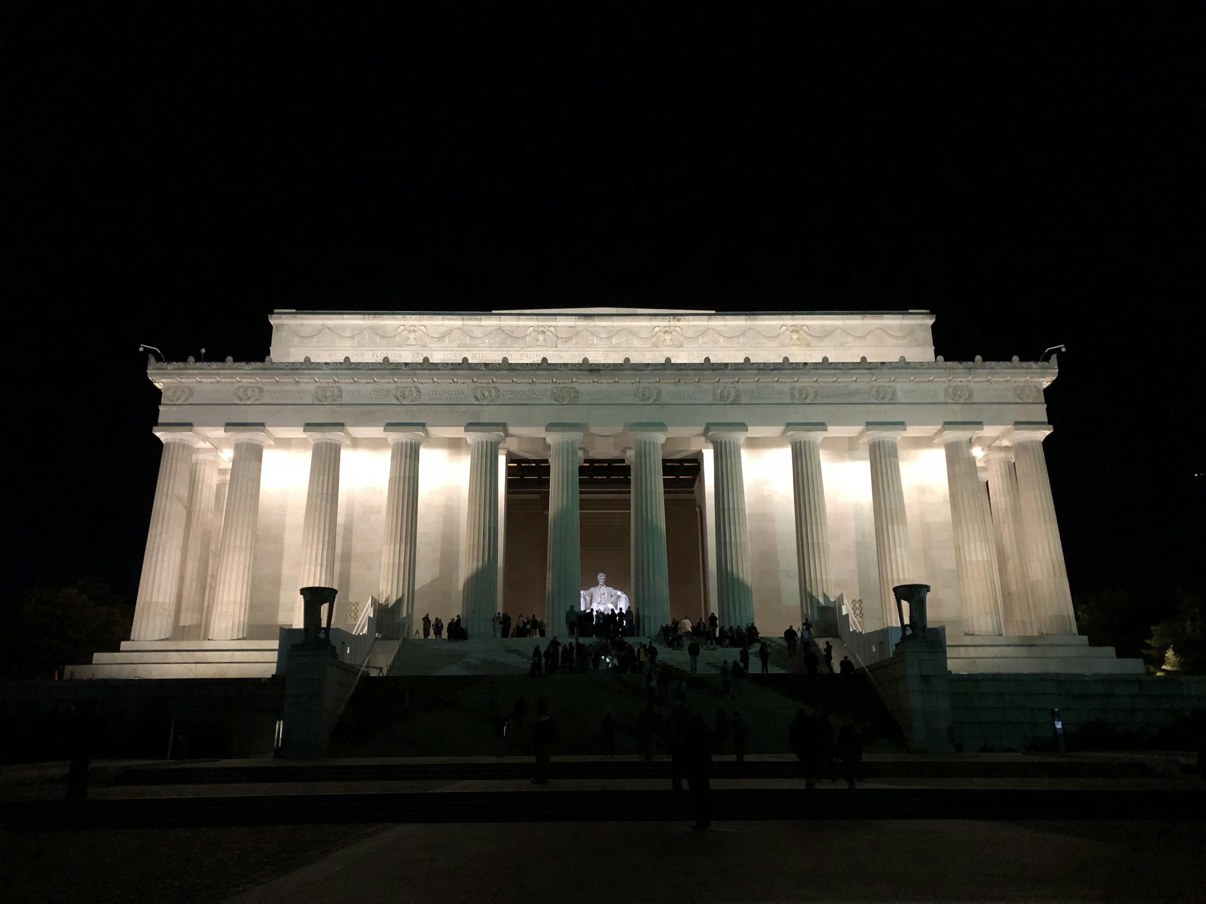 Lincoln Memorial bathed in soft light against a night sky, with visitors gathered at the base. The grandeur of the architecture is highlighted by the illumination.