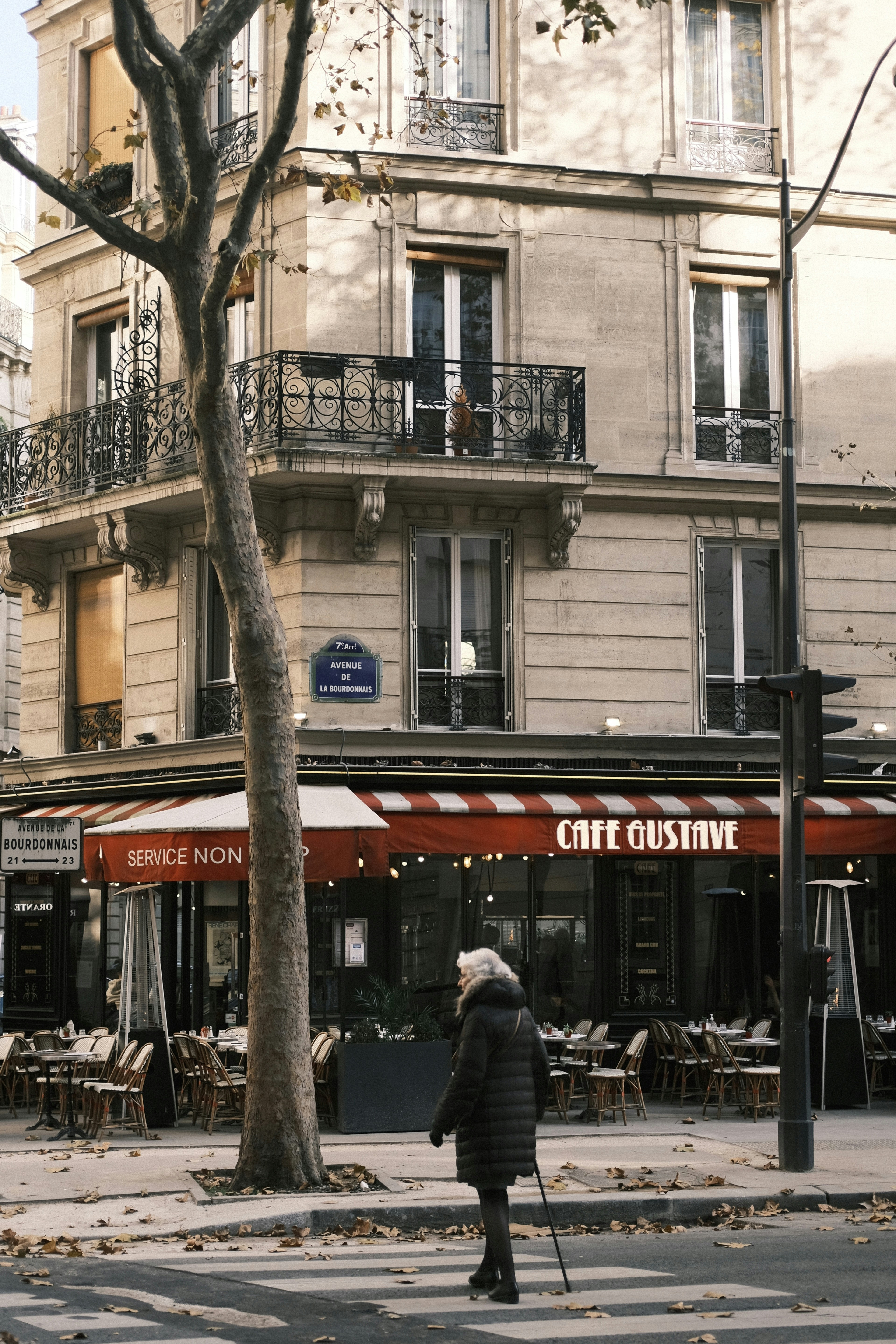 A woman walks past a quaint café with red awnings and outdoor seating, framed by elegant architecture in a bustling Paris street.