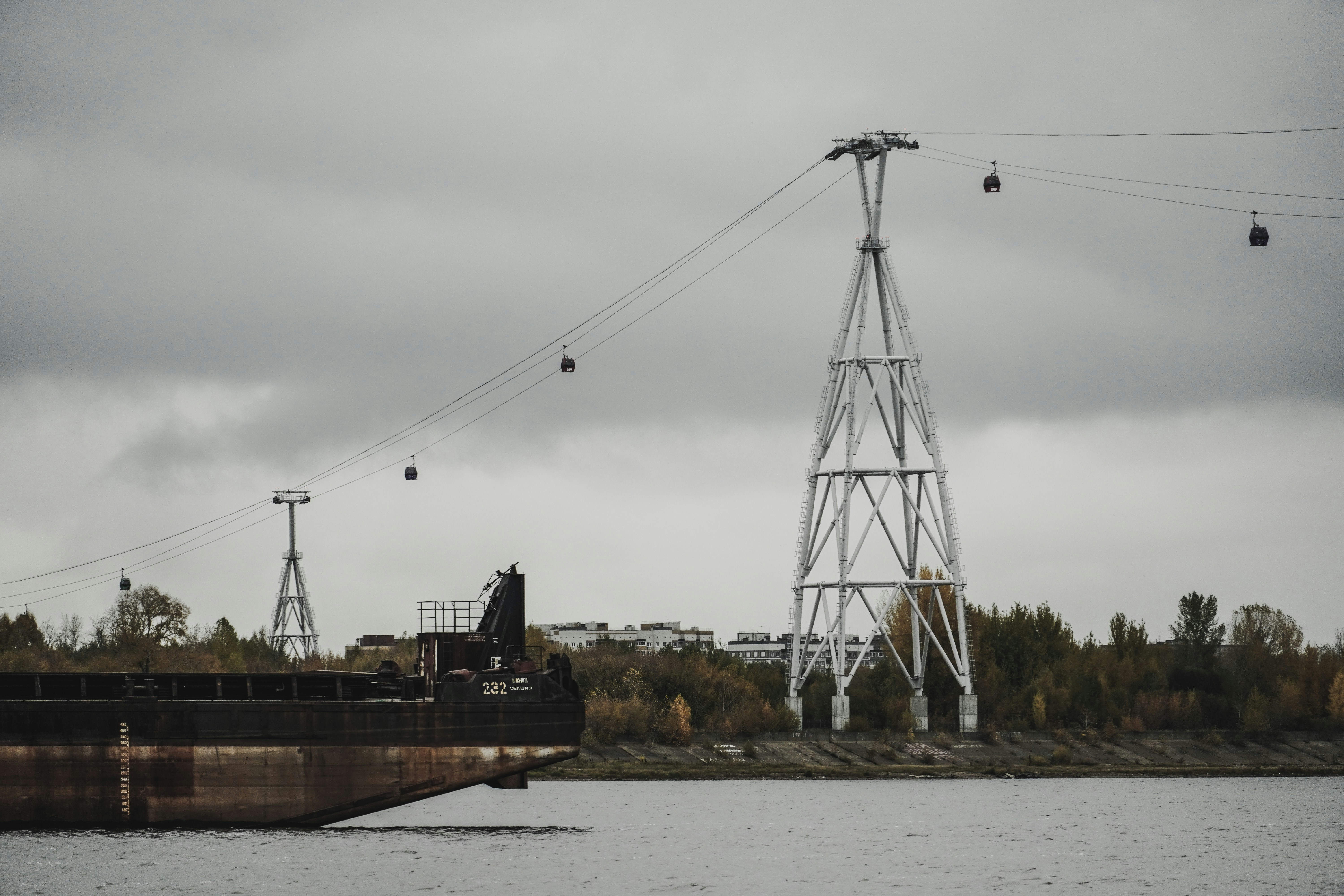 A barge navigates a river while a cable car system looms overhead, framed by a moody sky and autumn foliage. The industrial elements contrast with the natural landscape.