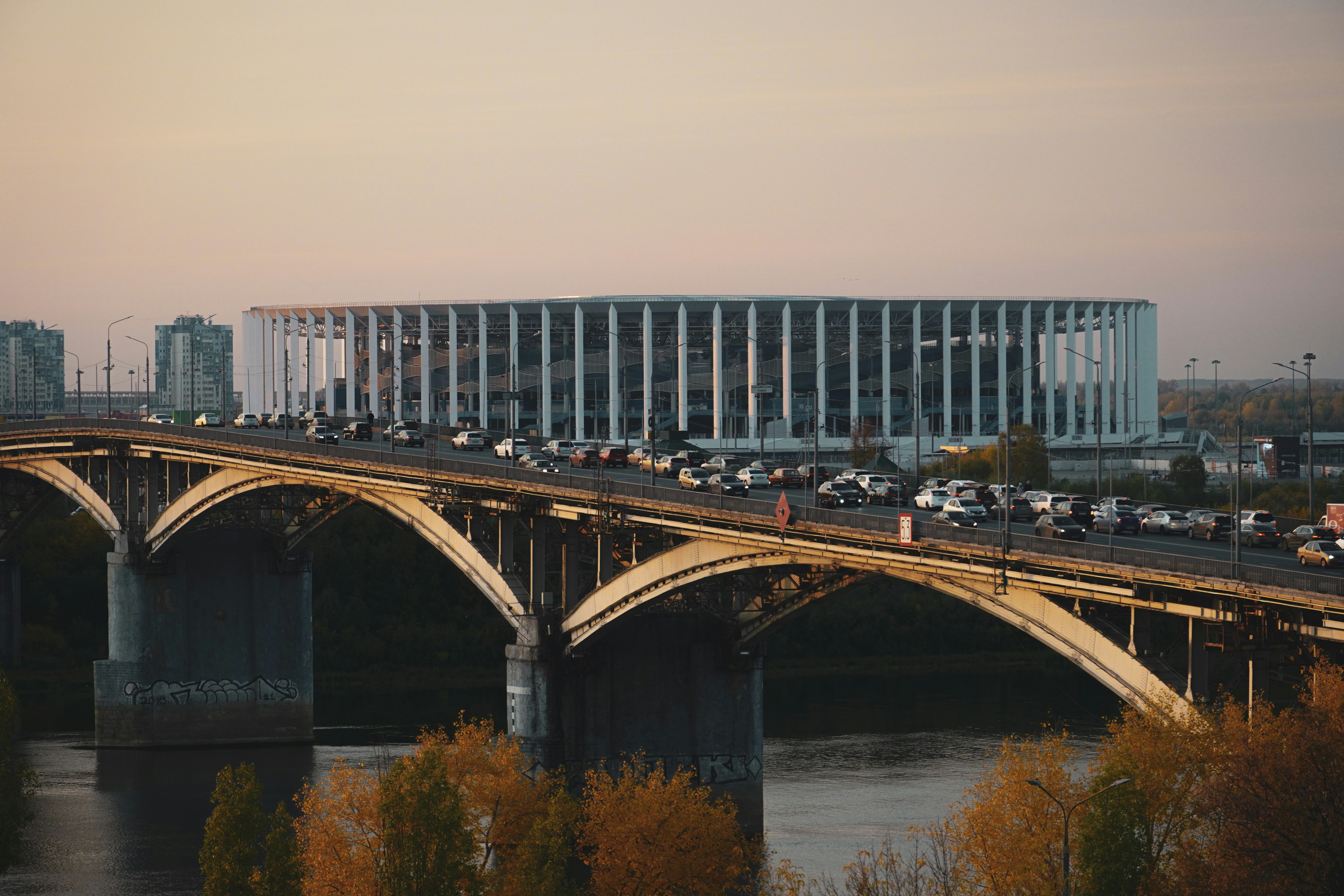 Cars traverse a busy bridge while a contemporary stadium looms in the background, framed by a soft evening sky.