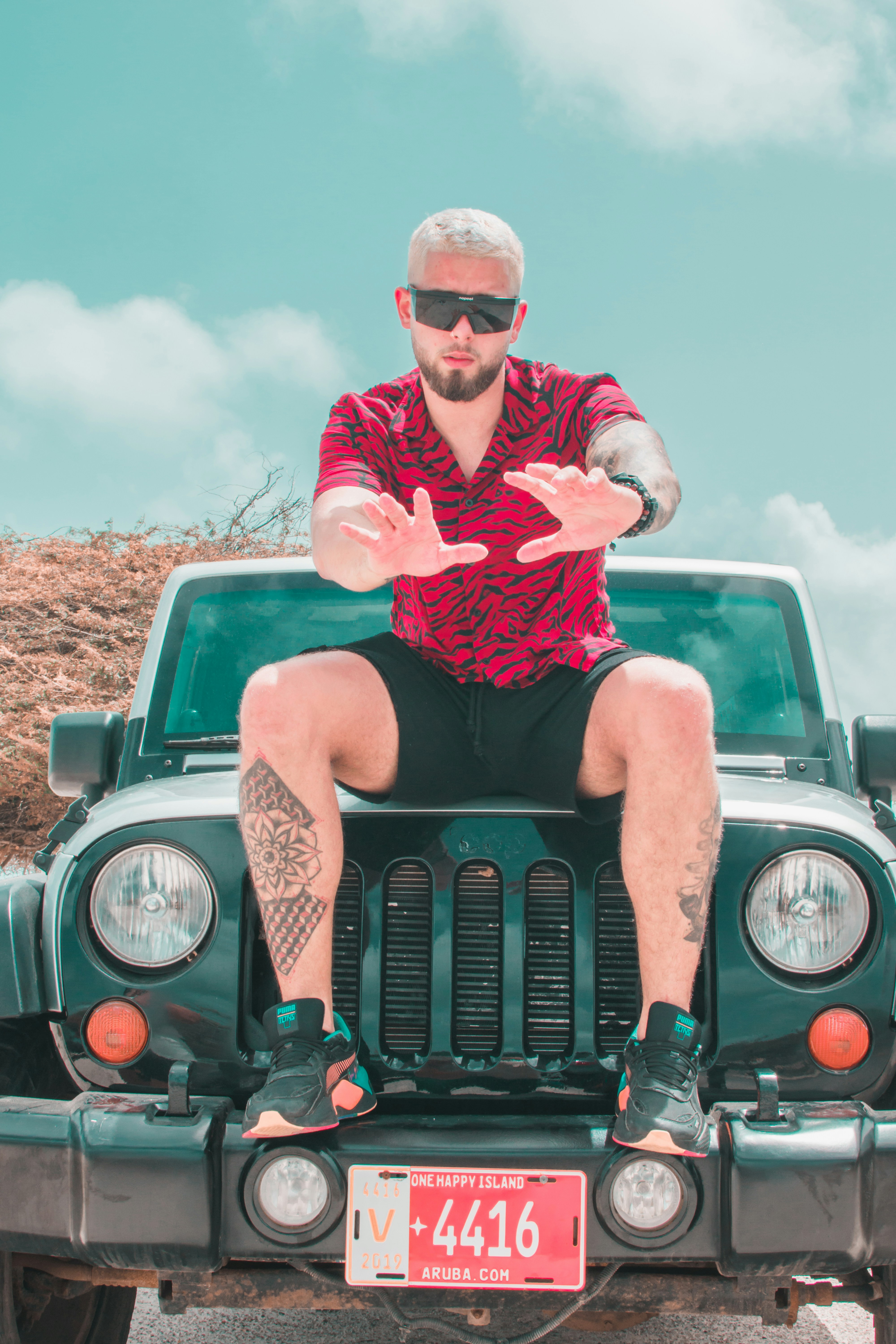 A man in a bold patterned shirt poses playfully on the hood of a black Jeep, set against a sunny backdrop with hints of nature. His confident stance and stylish sunglasses enhance the carefree atmosphere.