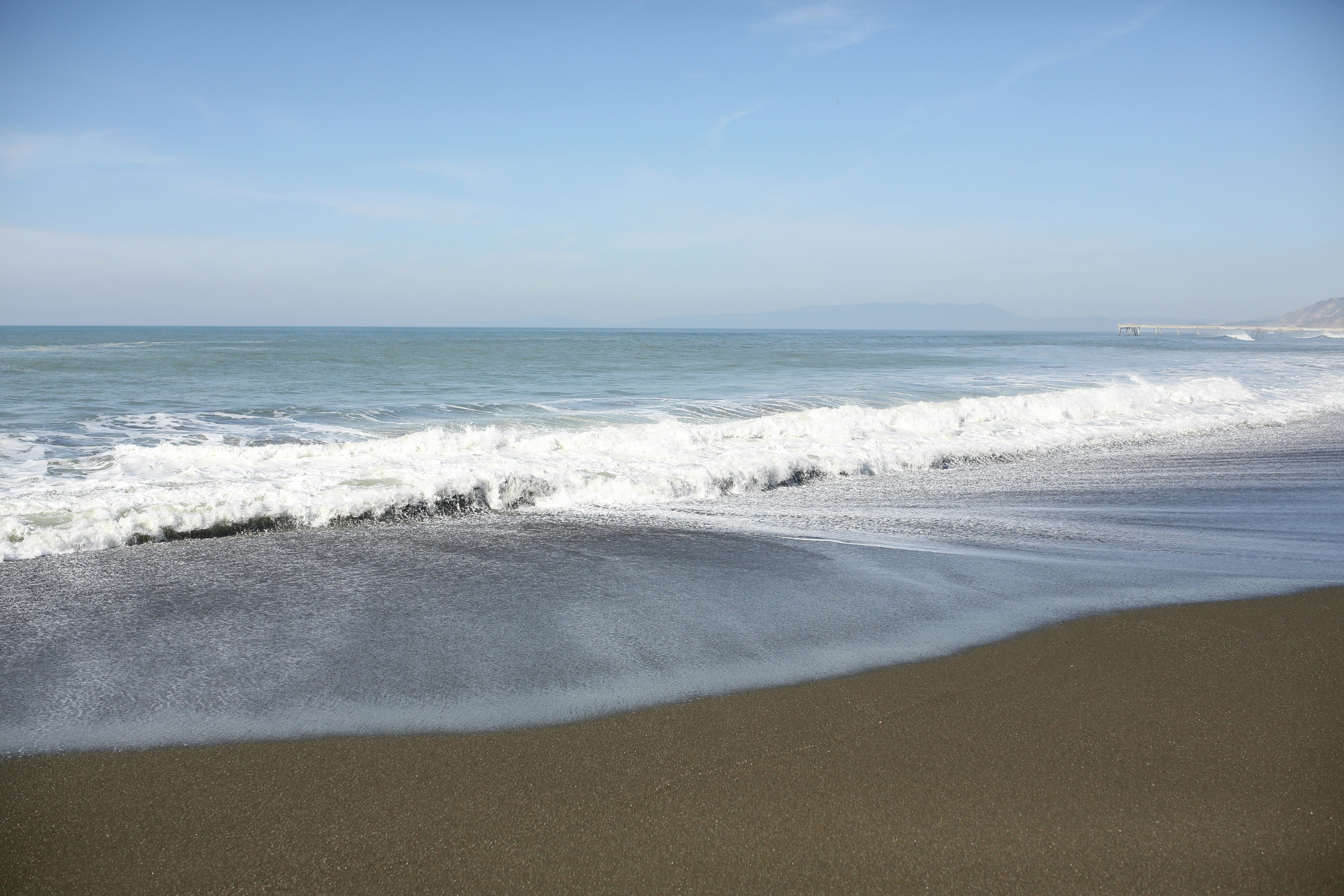 Gentle waves lapping against a dark sandy beach under a clear blue sky. The horizon stretches into the distance, merging sea and sky.