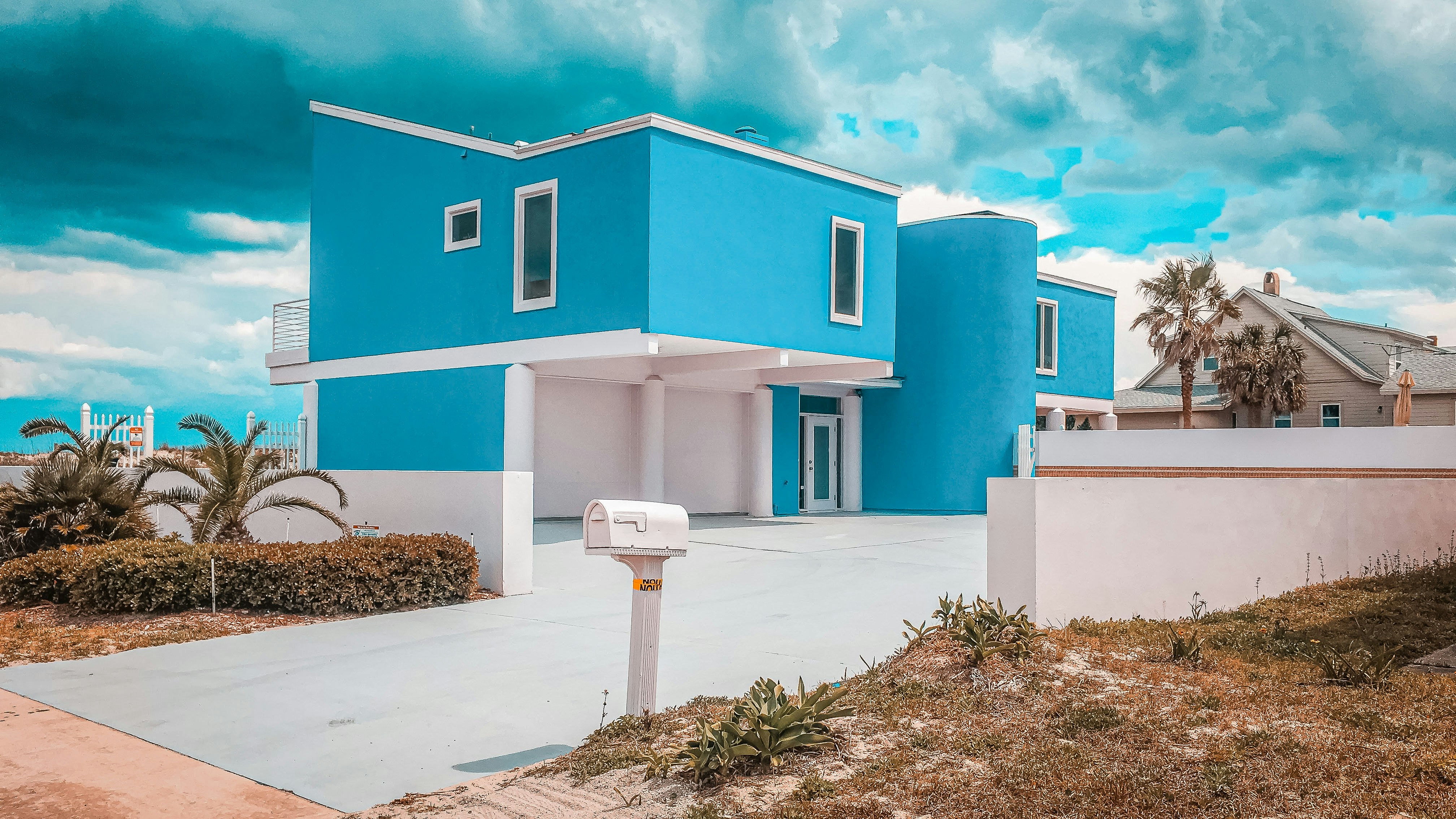 Vibrant blue contemporary house surrounded by greenery and palm trees, set against a dramatic sky.