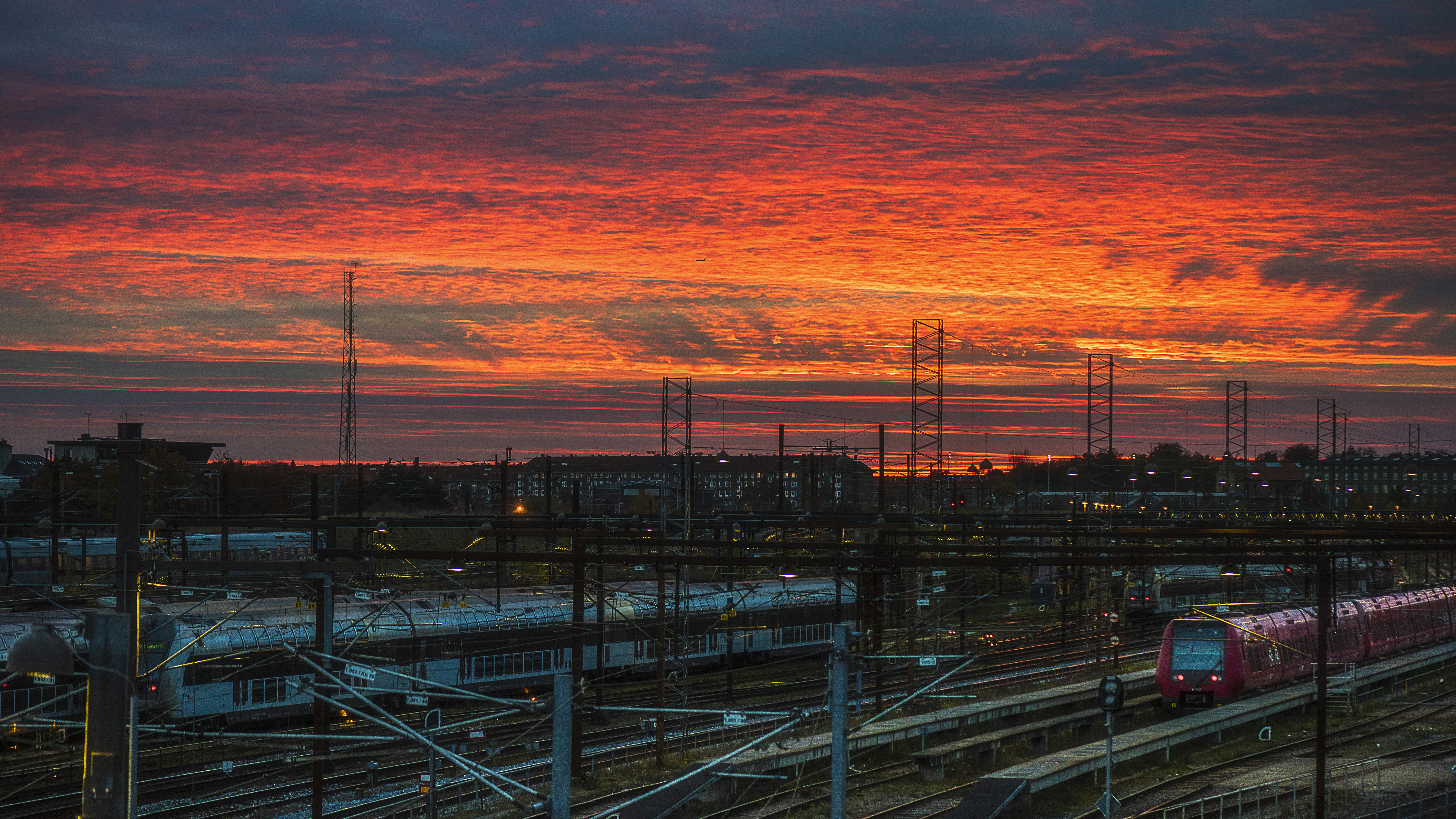 Vibrant sunset casts a dramatic glow over a bustling train yard, with tracks and trains silhouetted against the colorful sky.