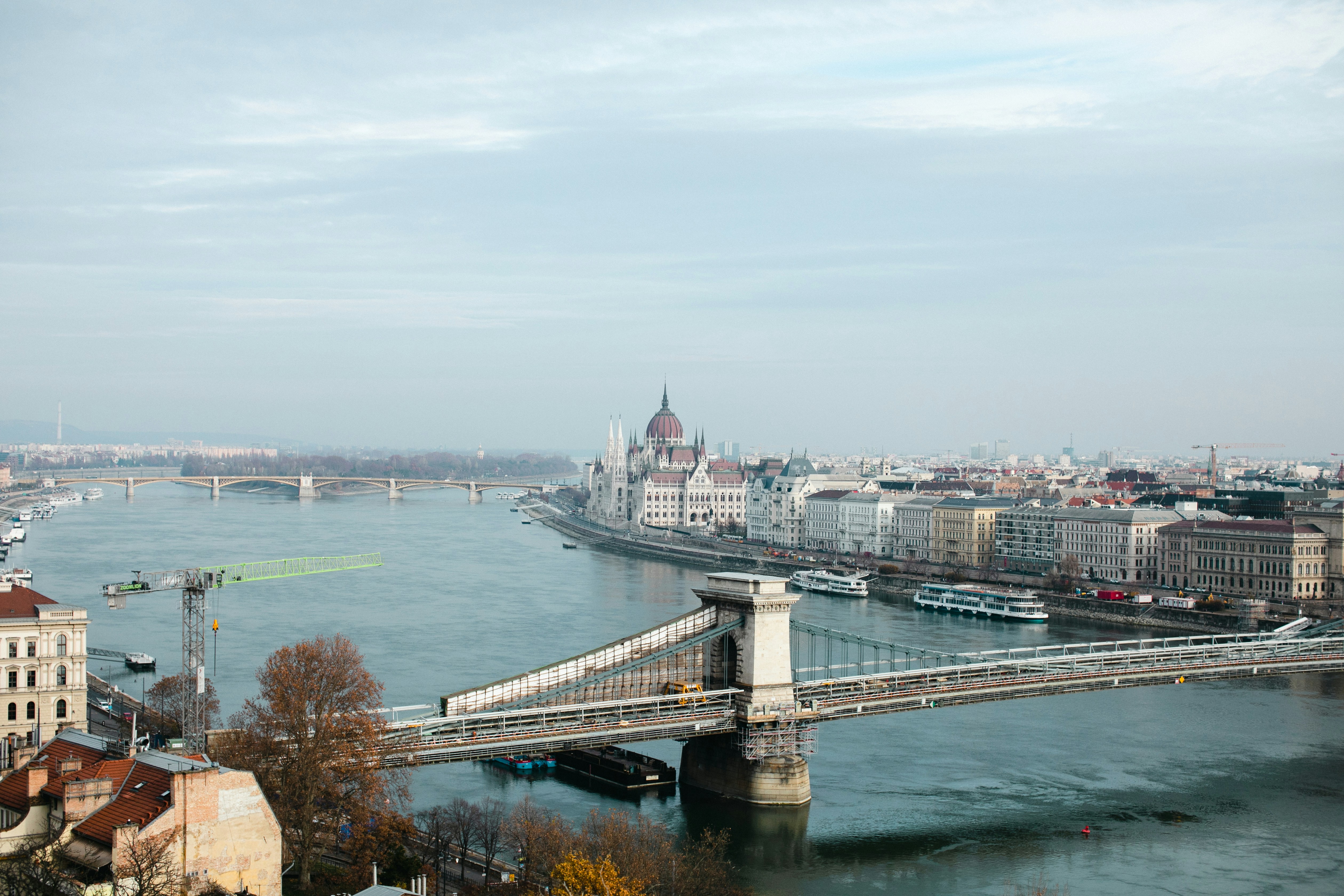 A panoramic view of the Danube River showcasing the iconic Chain Bridge and the Hungarian Parliament building in the background. The tranquil waters reflect the cityscape under a soft, cloudy sky.