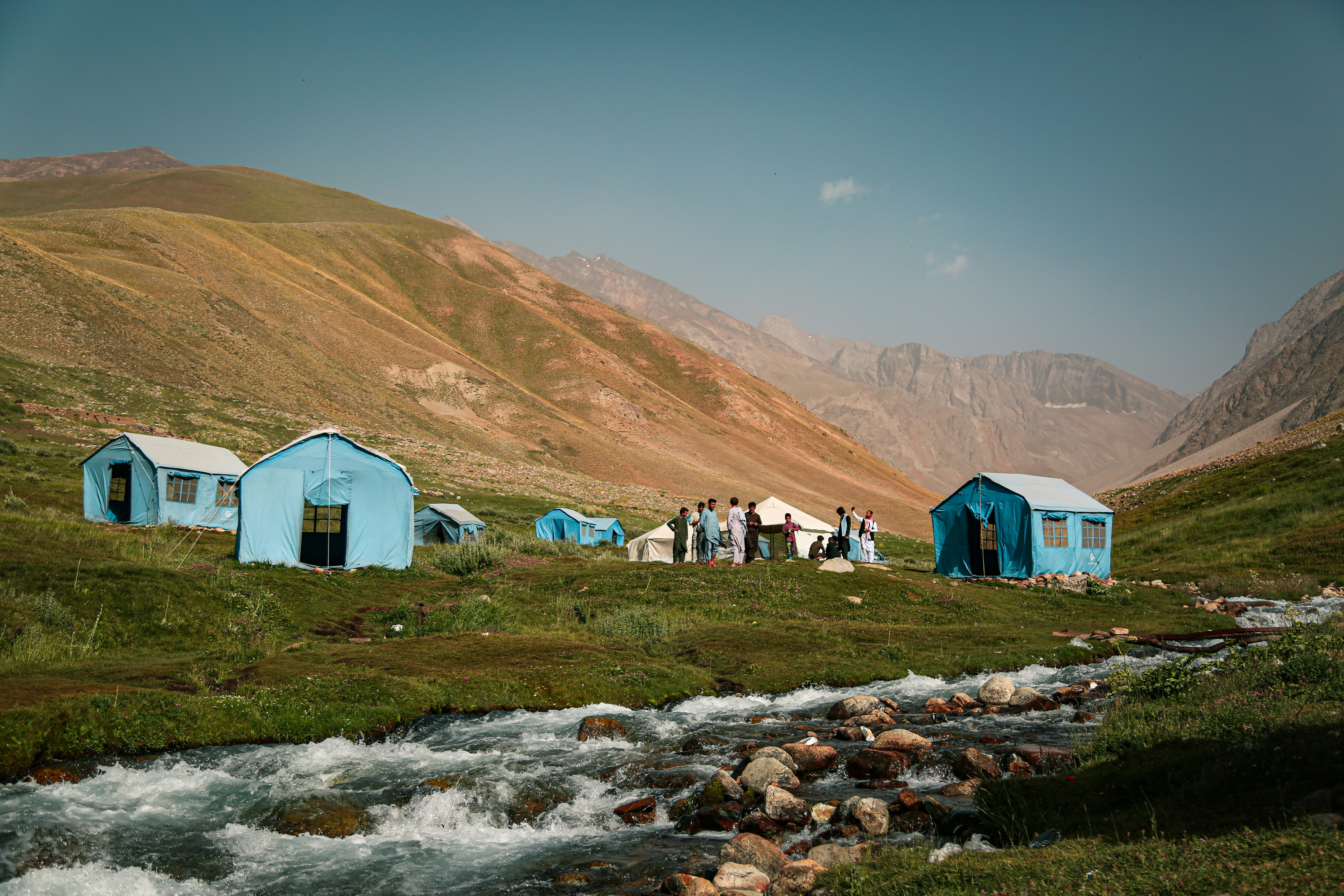Cluster of blue huts beside a clear stream in a vast, grassy mountain valley under a clear sky.