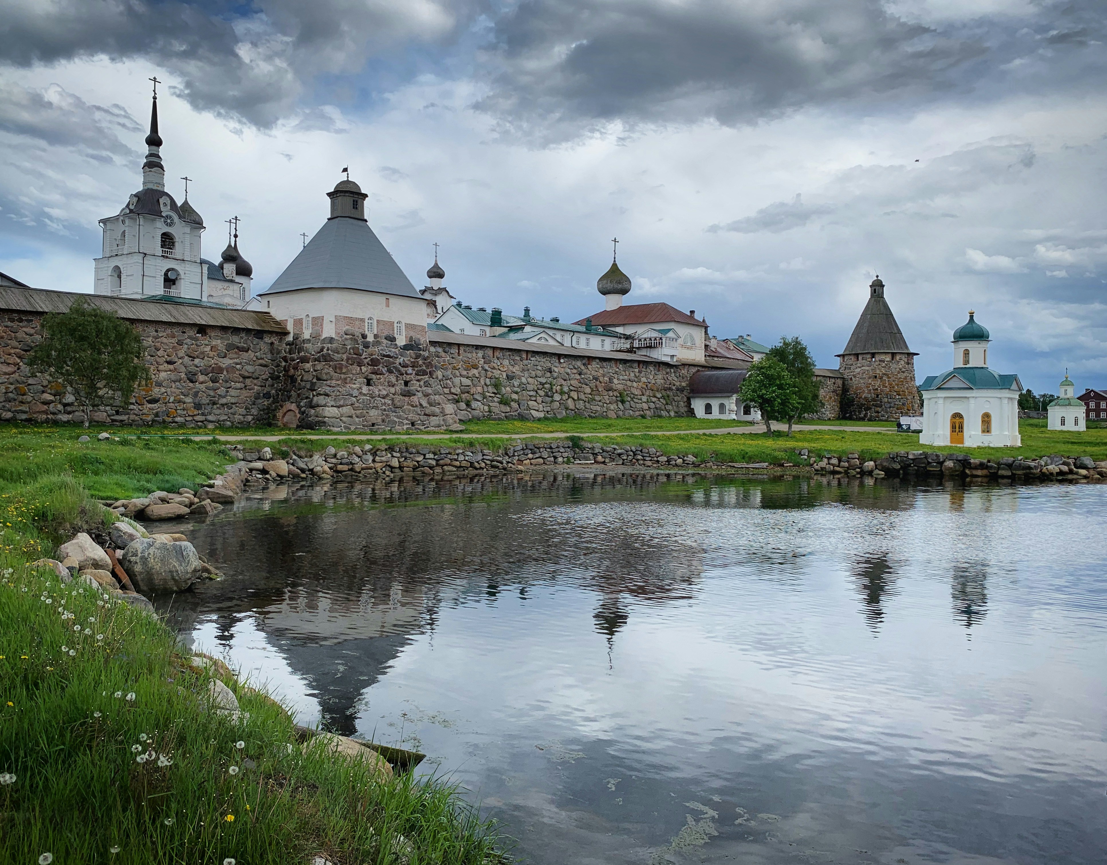 Historic monastery surrounded by a tranquil pond, with stone walls and various architectural features reflecting in the water.