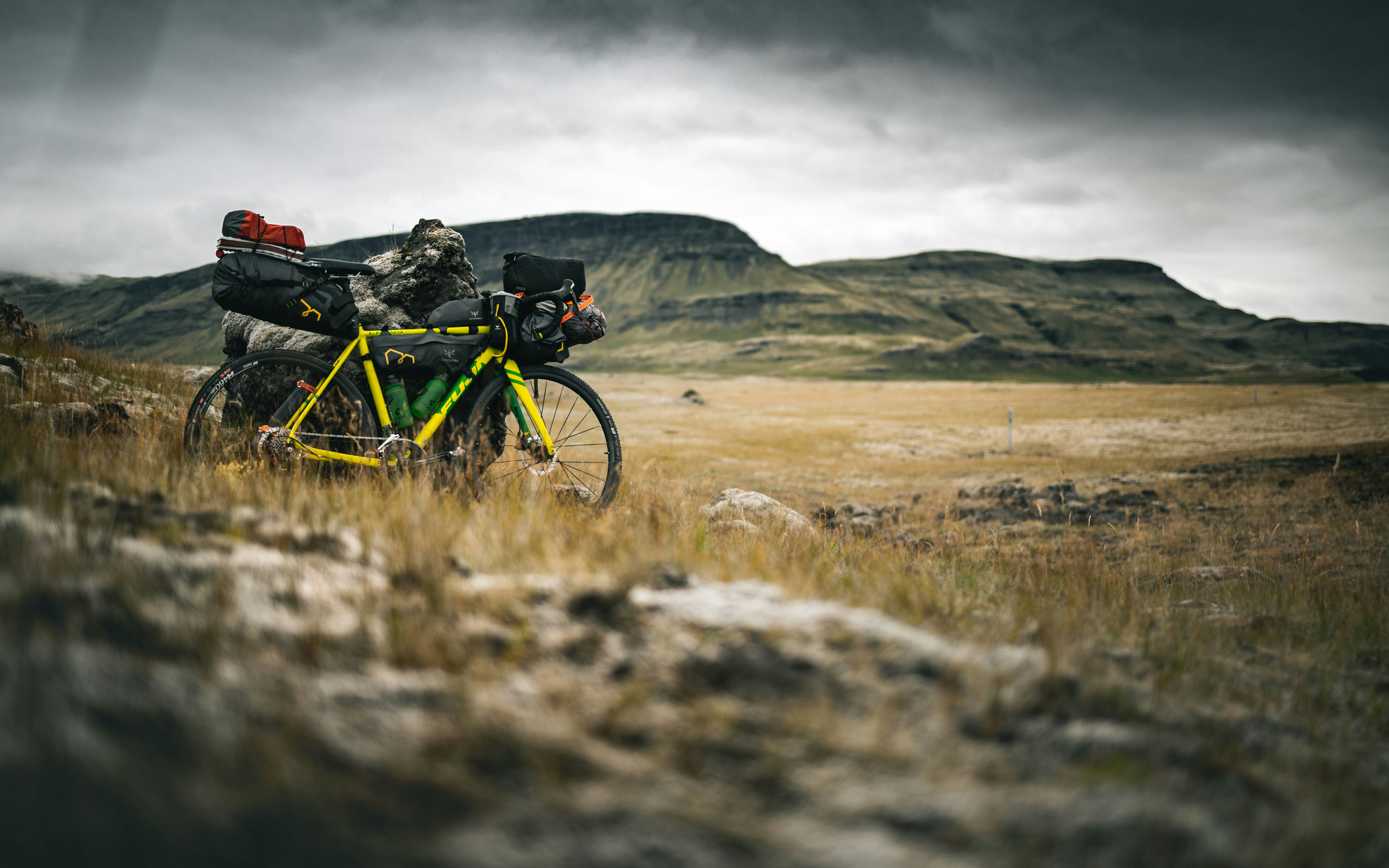 A brightly colored bicycle equipped for touring rests against rocky terrain, set against rolling hills under an overcast sky.