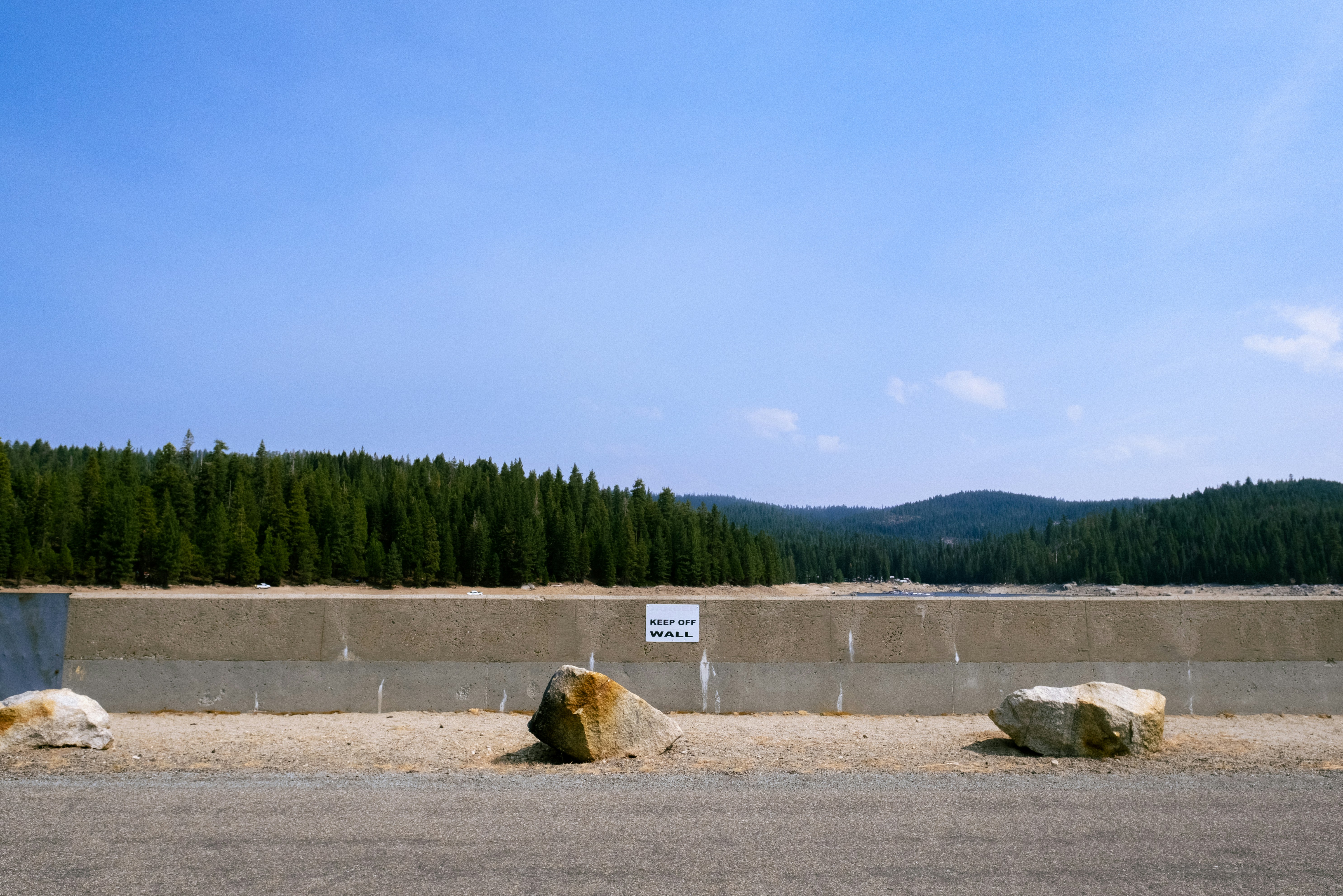 Concrete barrier with large rocks in the foreground, set against a forest and distant hills under a clear blue sky.