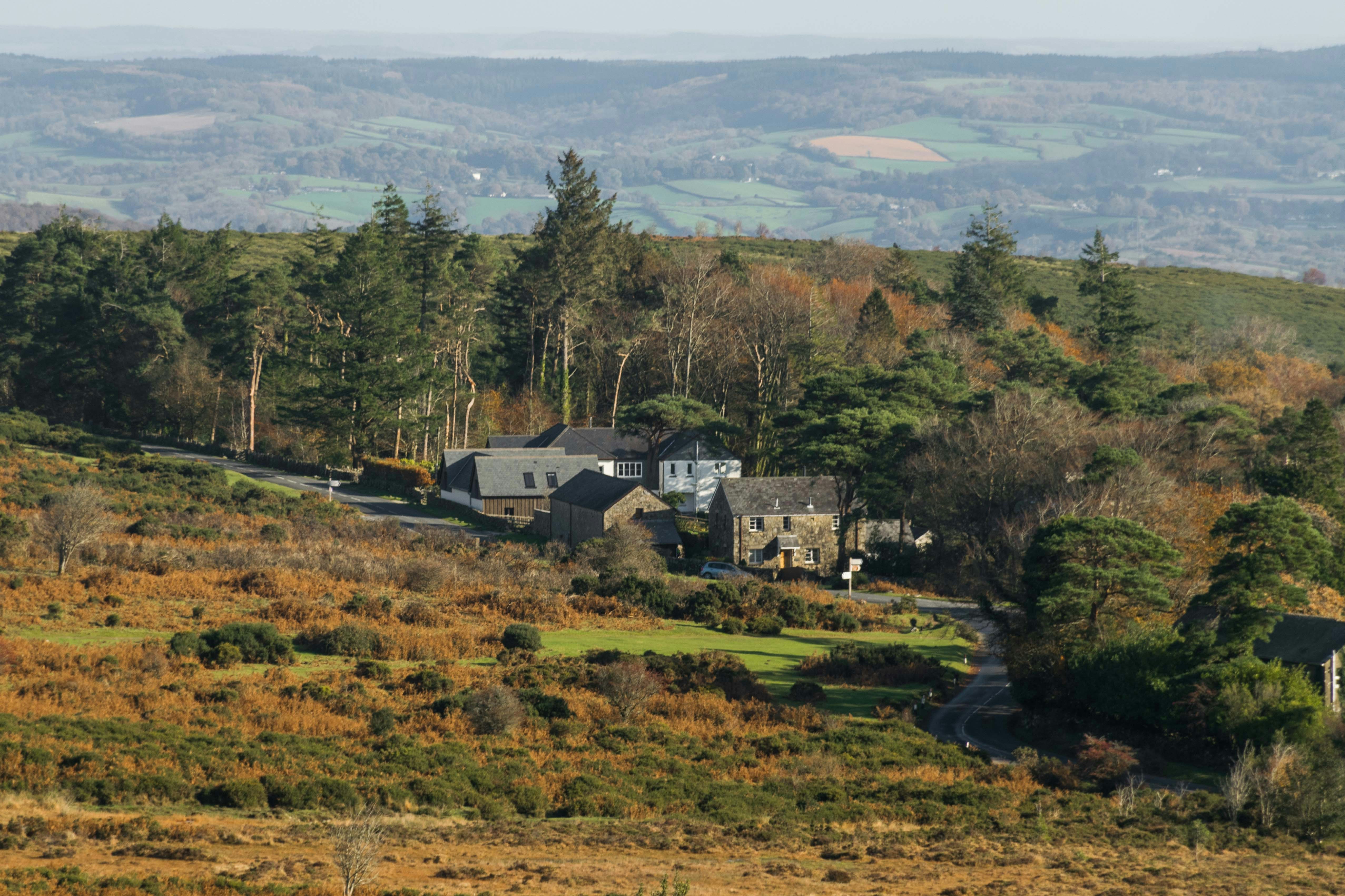 Charming cluster of rustic buildings nestled in a lush, hilly landscape, surrounded by autumn foliage and distant hills. The scene conveys a peaceful rural atmosphere.
