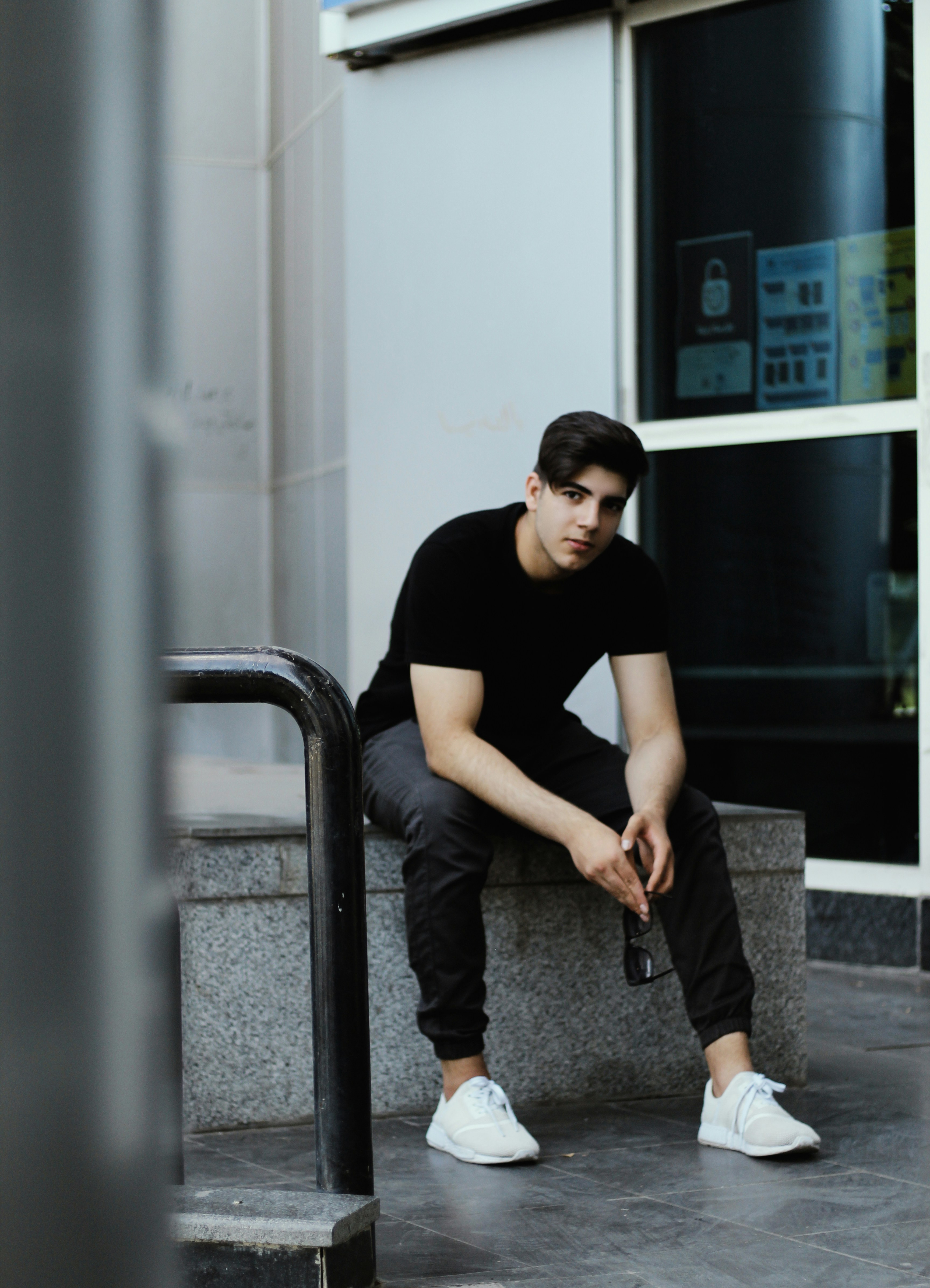 A young man dressed in black sits on a stone step, holding a phone, with modern architecture in the background.
