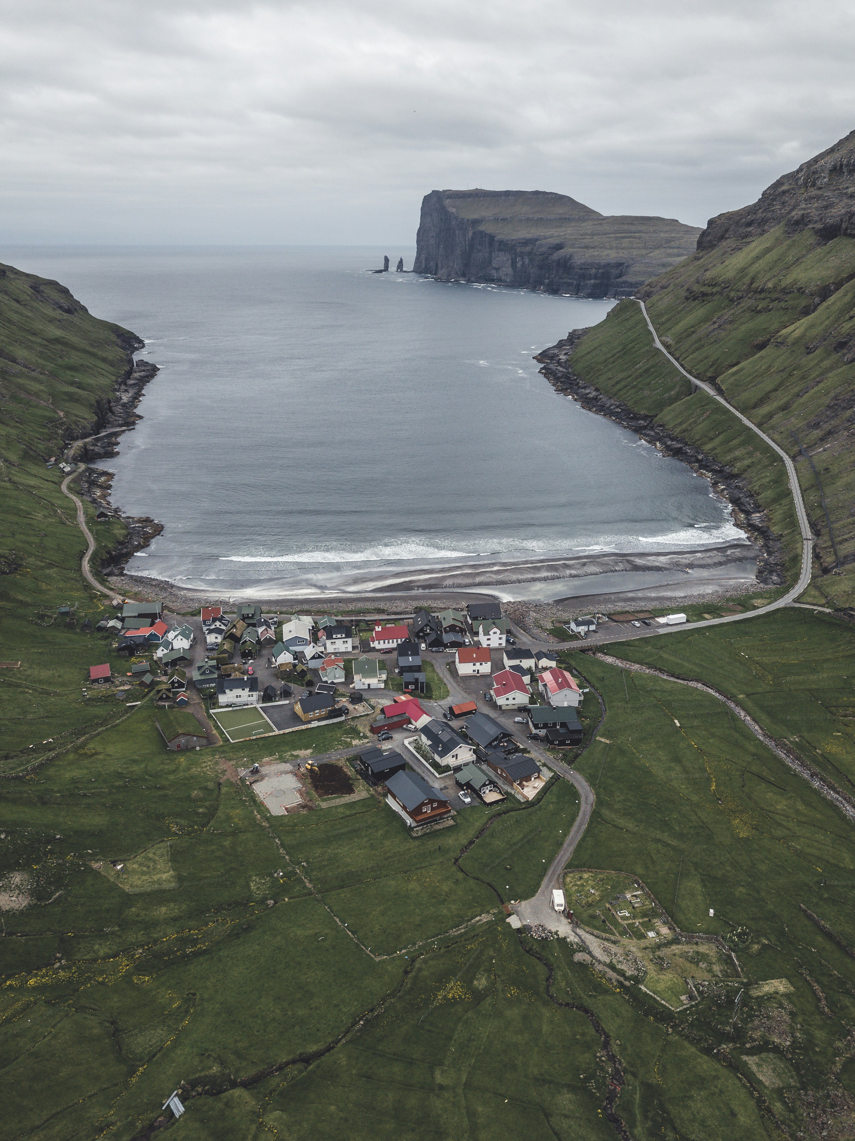 Aerial view of a quaint coastal village nestled between lush green hills and a tranquil sea, with dramatic cliffs in the distance. The scene highlights the harmony between human habitation and natural beauty.