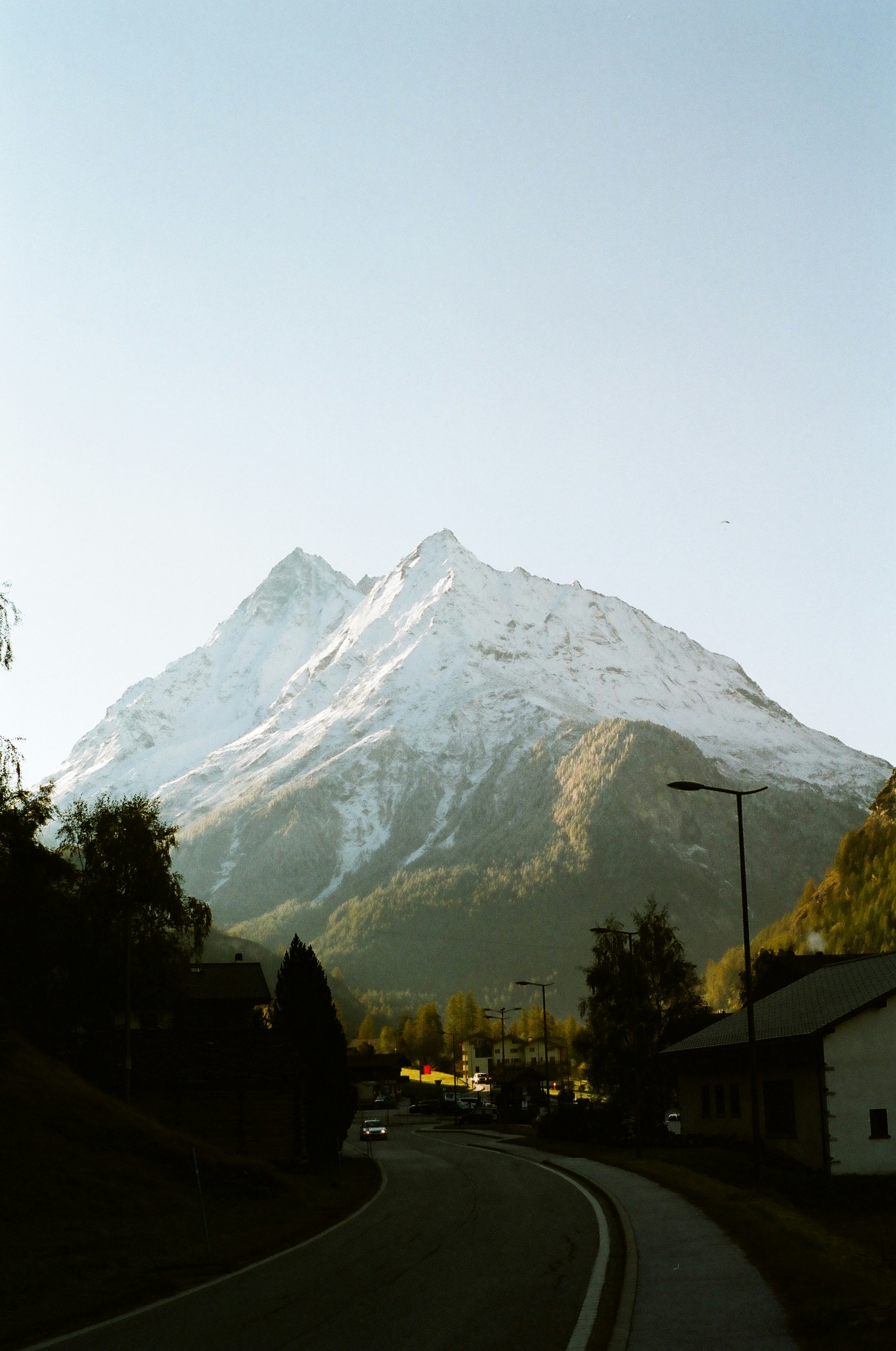 Snow-capped mountain dominates the scene, with a winding road and a small village at its base beneath a clear blue sky.