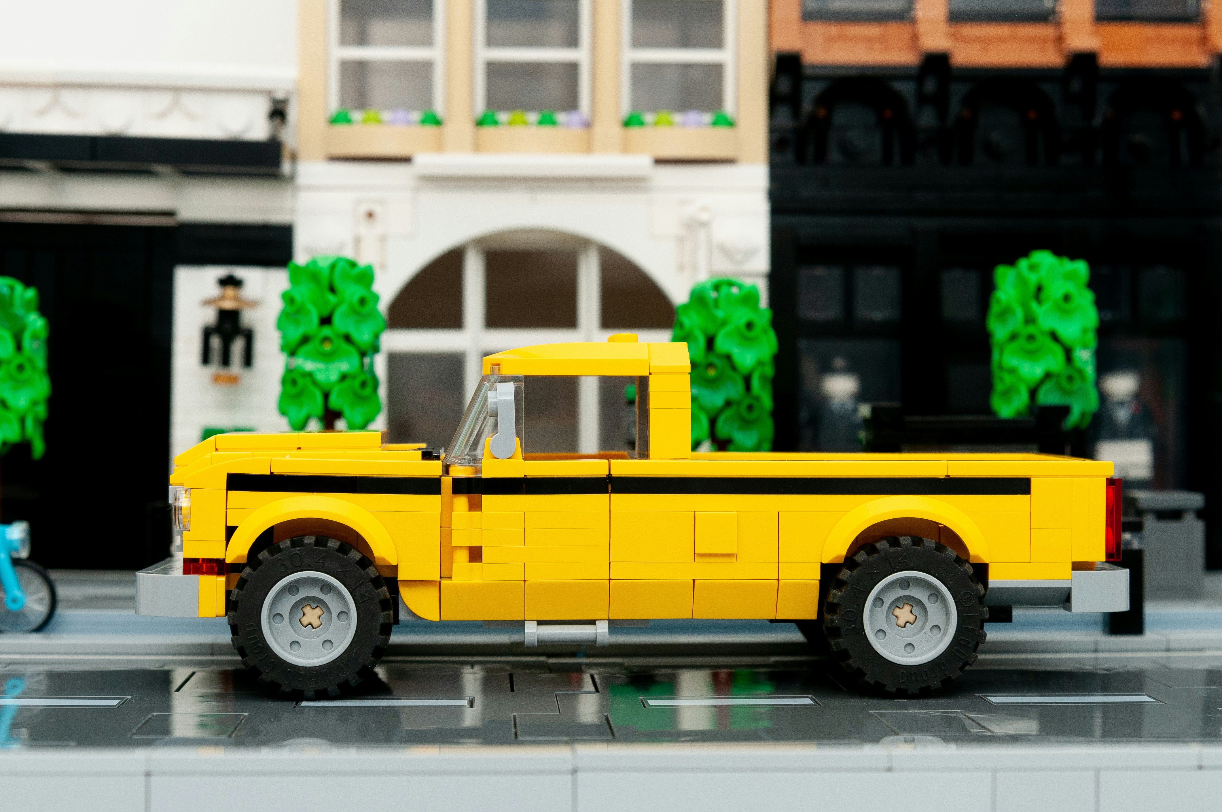 A bright yellow Lego pickup truck parked on a city street, surrounded by lush green foliage and urban architecture.