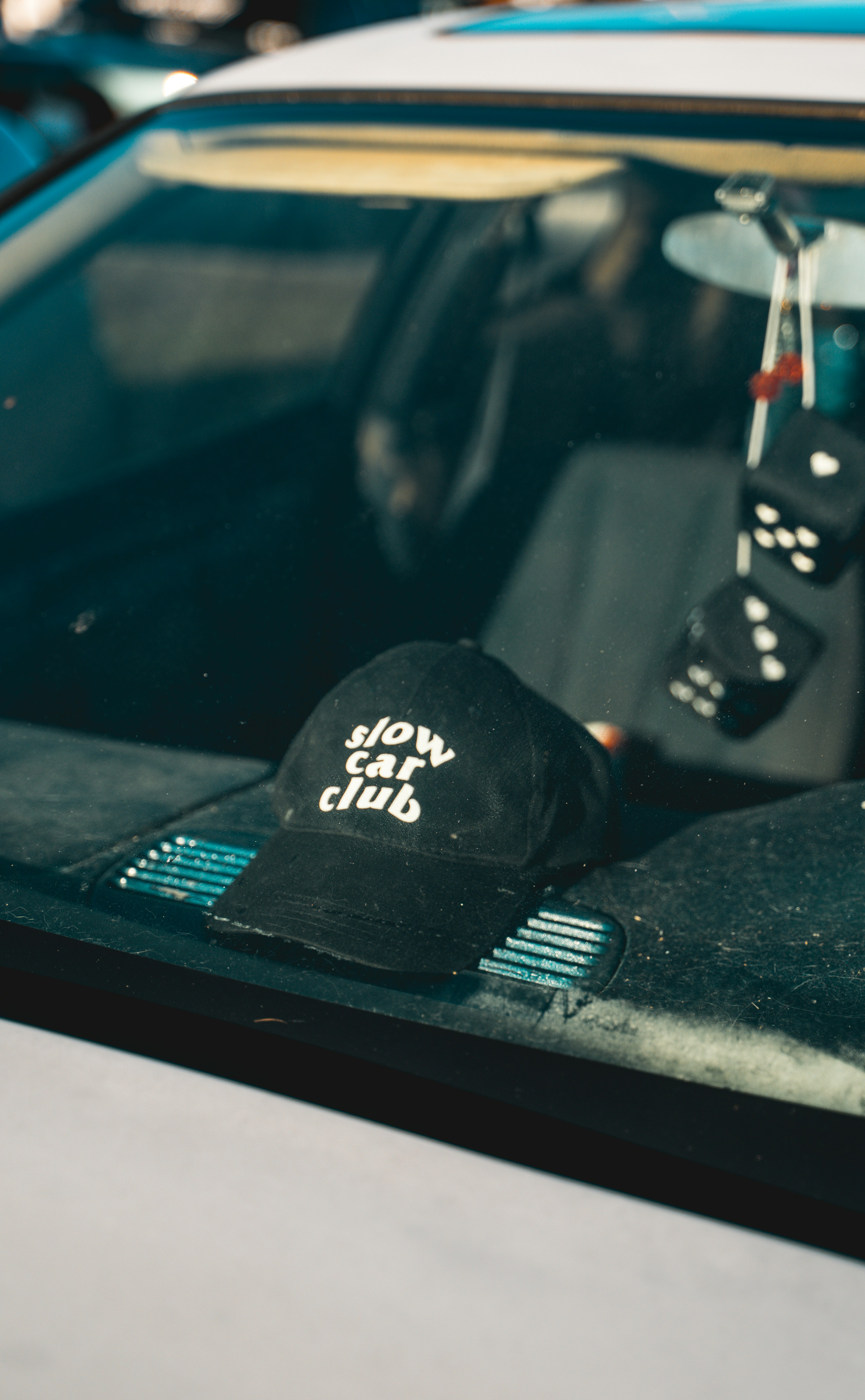 A black cap with 'slow car club' embroidered on it rests on the dashboard of a vintage car, hinting at a laid-back lifestyle. The scene captures a moment of casual charm.