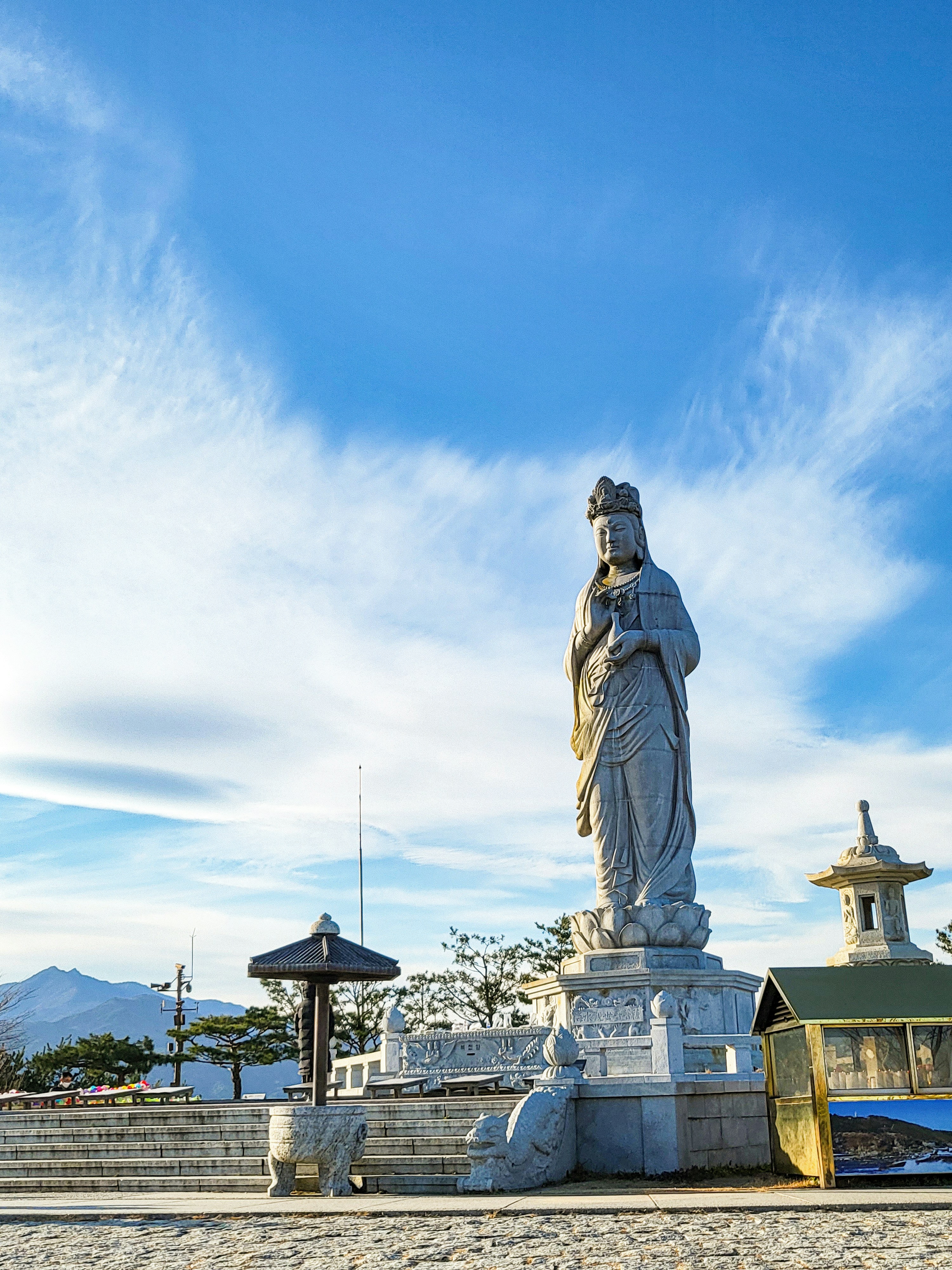 Majestic statue of a serene figure standing against a backdrop of blue sky and scattered clouds, surrounded by traditional architecture and nature.