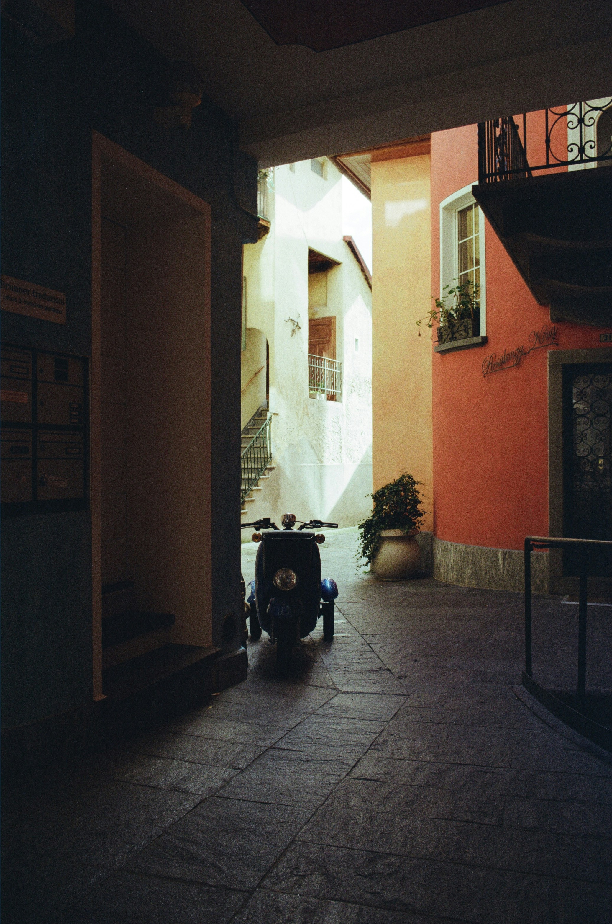 A quaint alleyway featuring a vintage scooter parked at the entrance, framed by colorful buildings and lush greenery.
