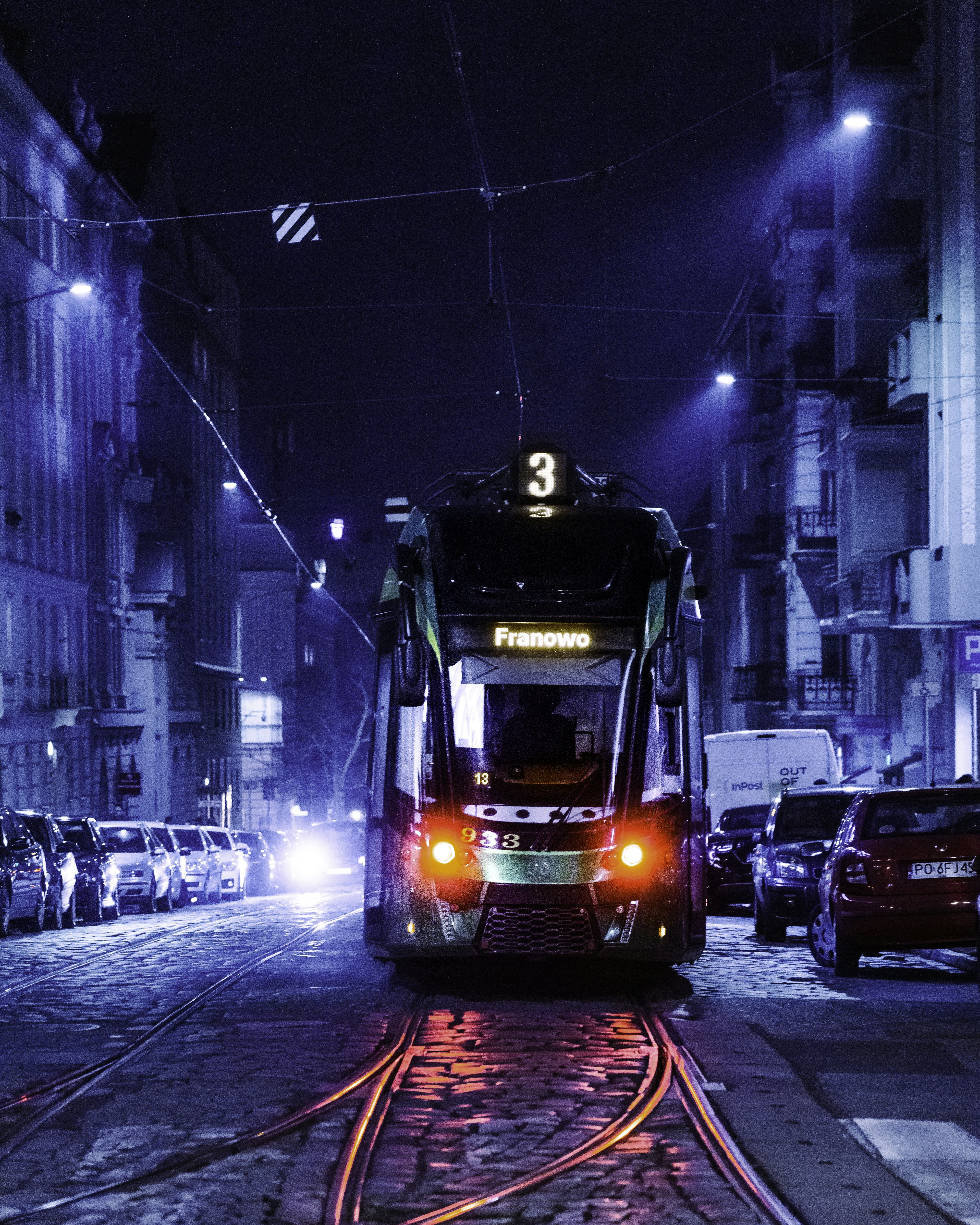 A tram labeled 'Franowo' navigates a dimly lit street, flanked by parked cars, under the glow of streetlights. The scene captures the essence of urban nightlife.