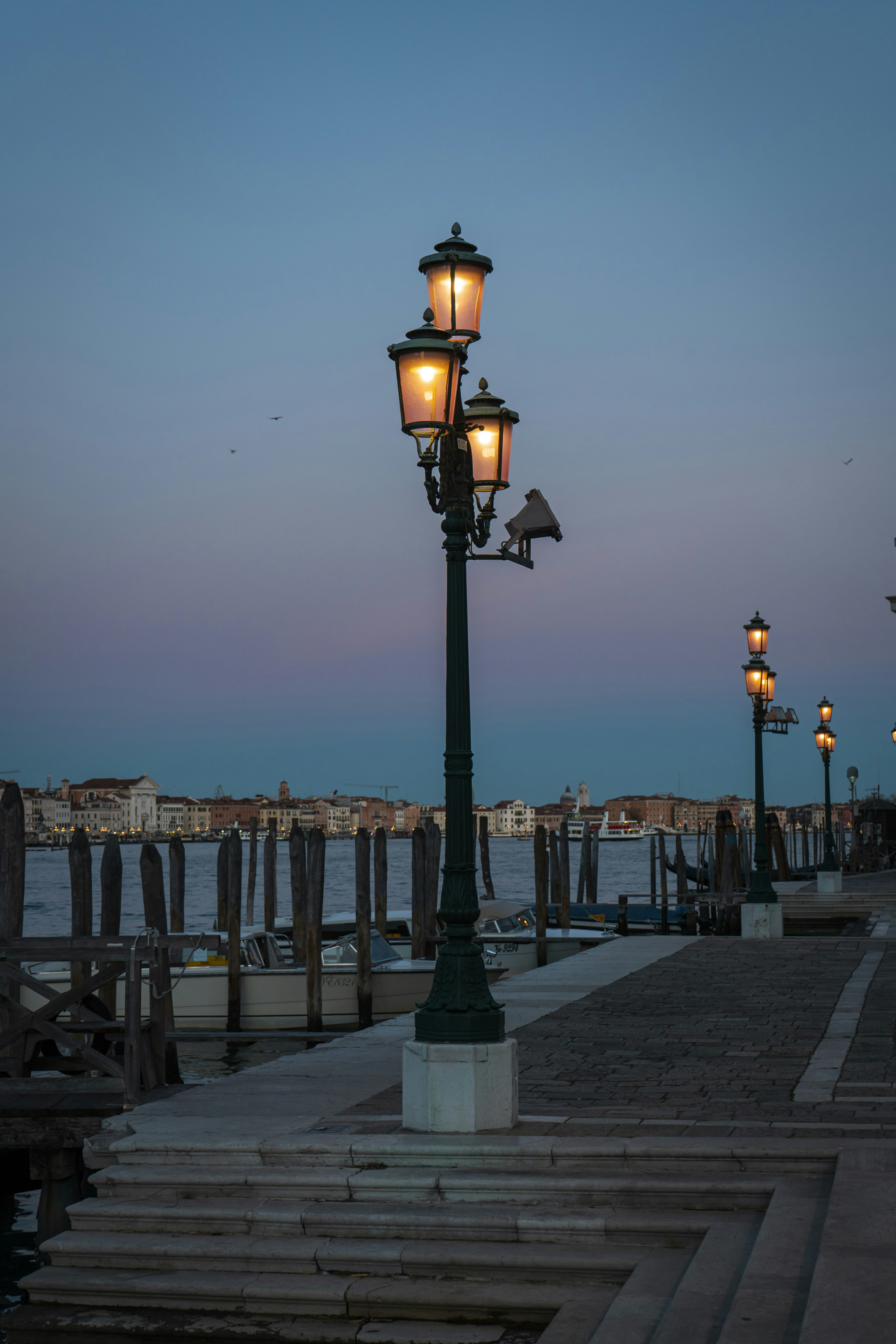 Elegant street lamps illuminate a quiet Venetian promenade at dusk, with a serene canal and distant cityscape in the background.
