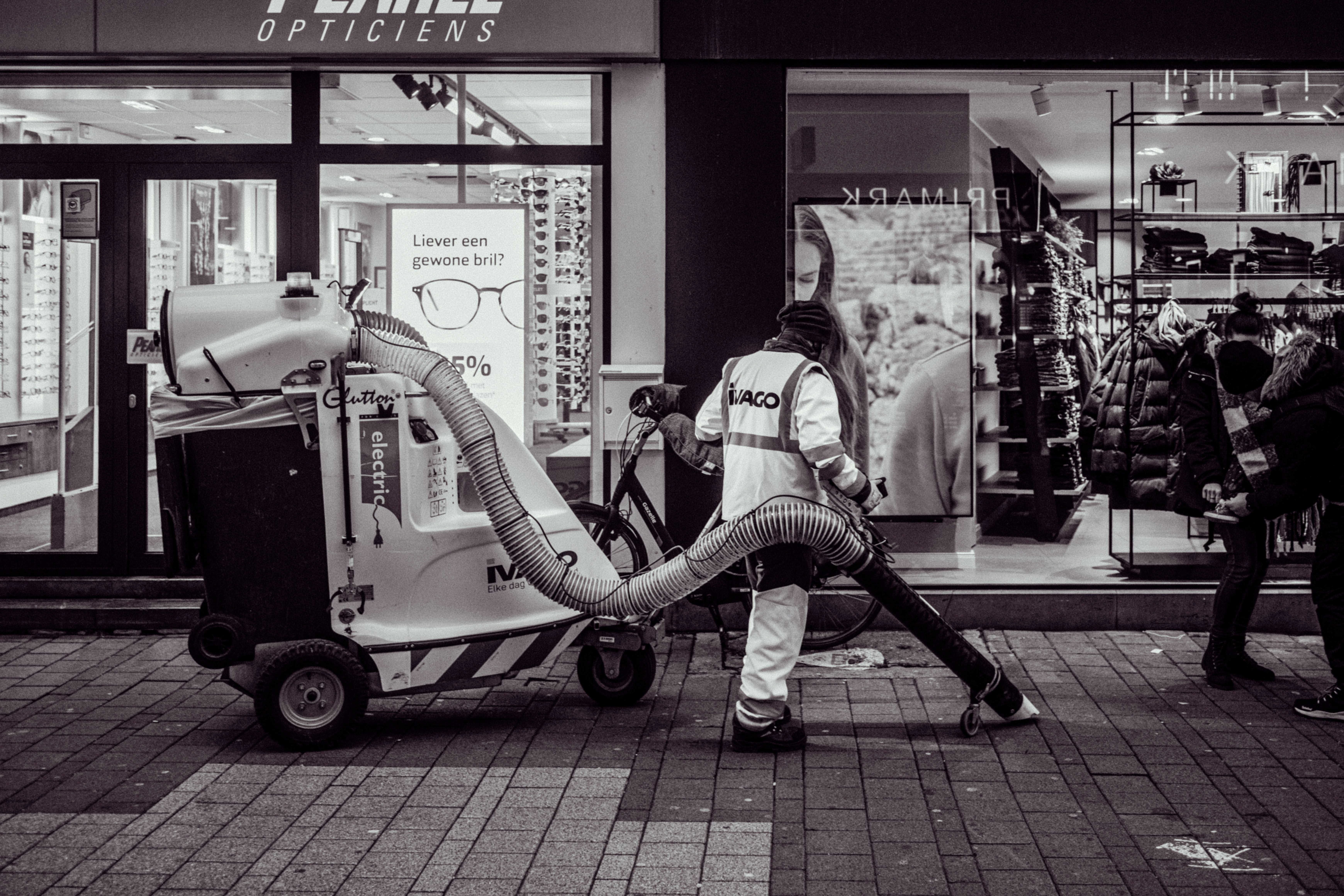 Hose and Haze: A wheeled extractor snakes past shopfronts in a grayscale city