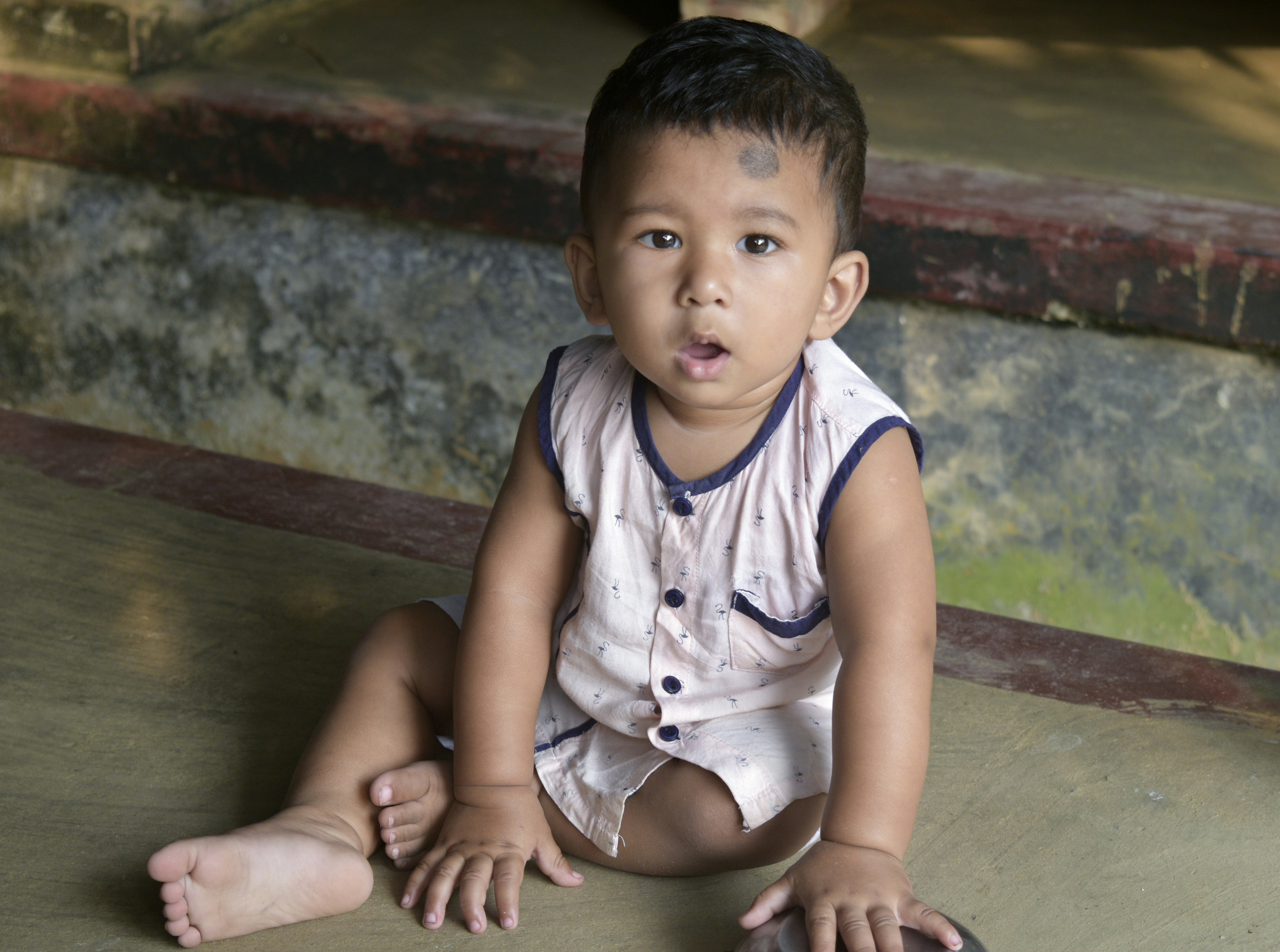 A young child sits on a rustic floor, gazing curiously at the viewer, with a playful expression and a hint of mischief. The setting features warm tones and natural textures.