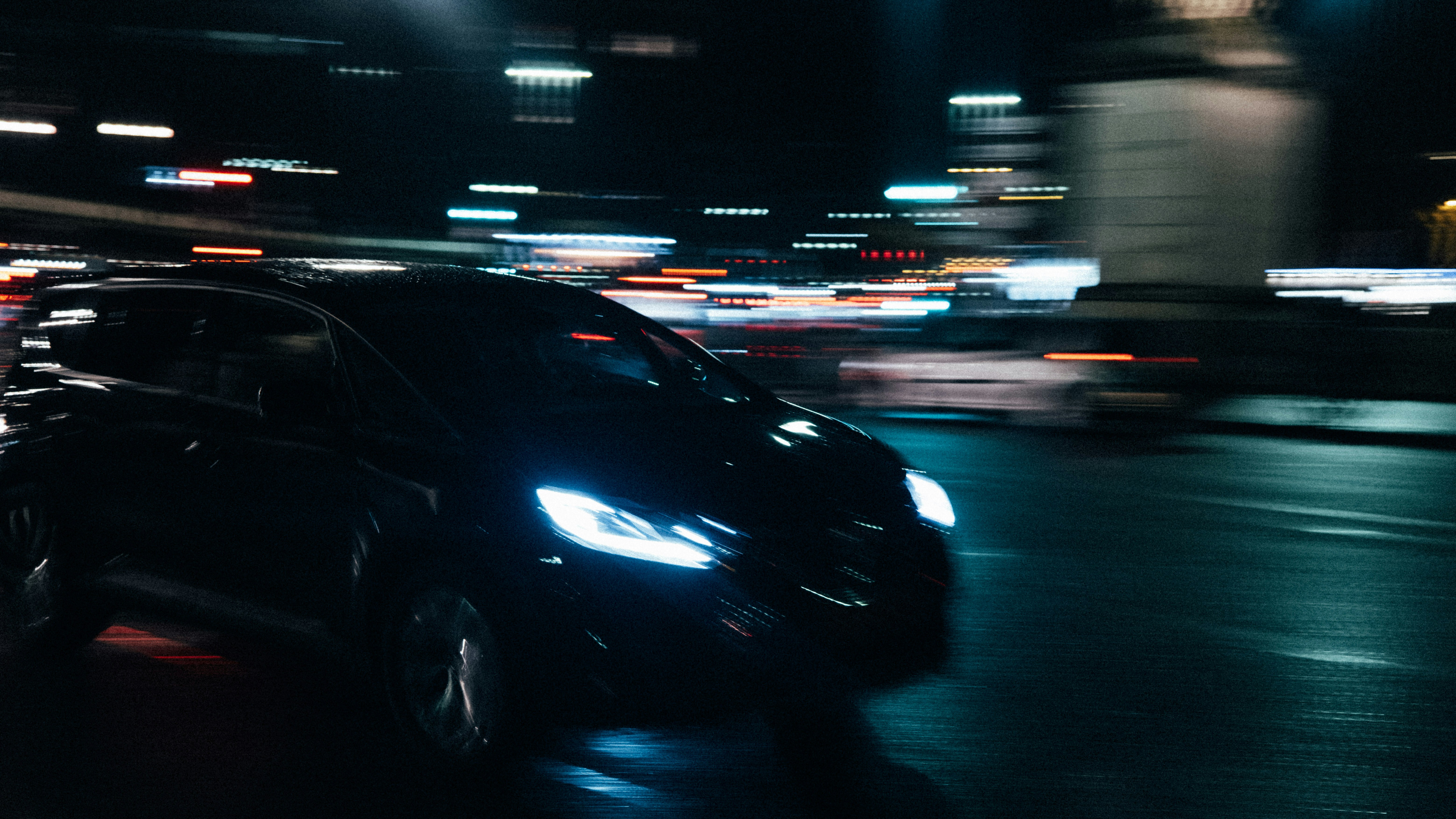 A sleek black car speeds through a vibrant cityscape at night, with streaks of colorful lights illuminating the wet pavement.