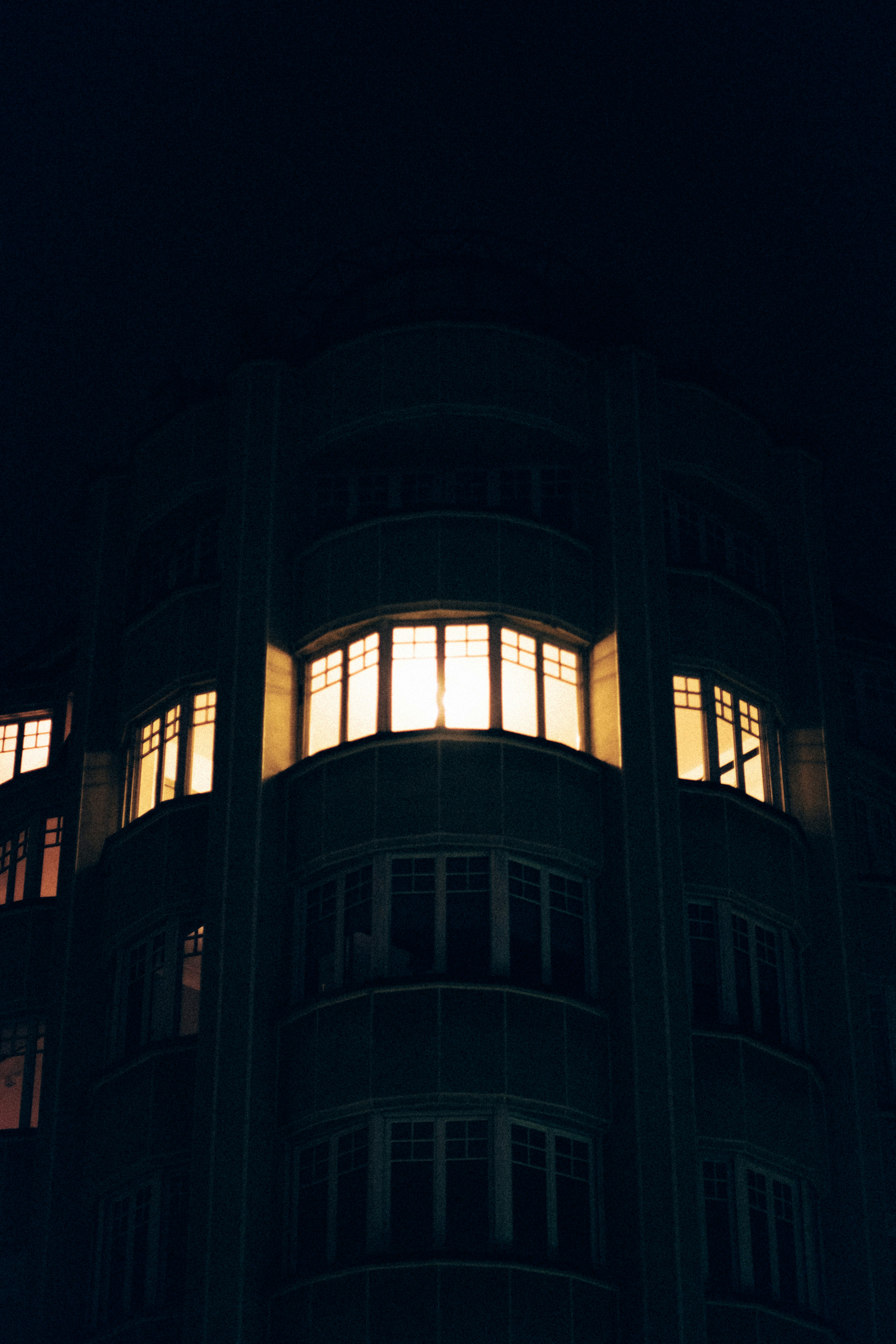 Warm light spills from a window of an art deco building at night, contrasting with the dark exterior. The scene evokes a sense of mystery and quiet reflection.