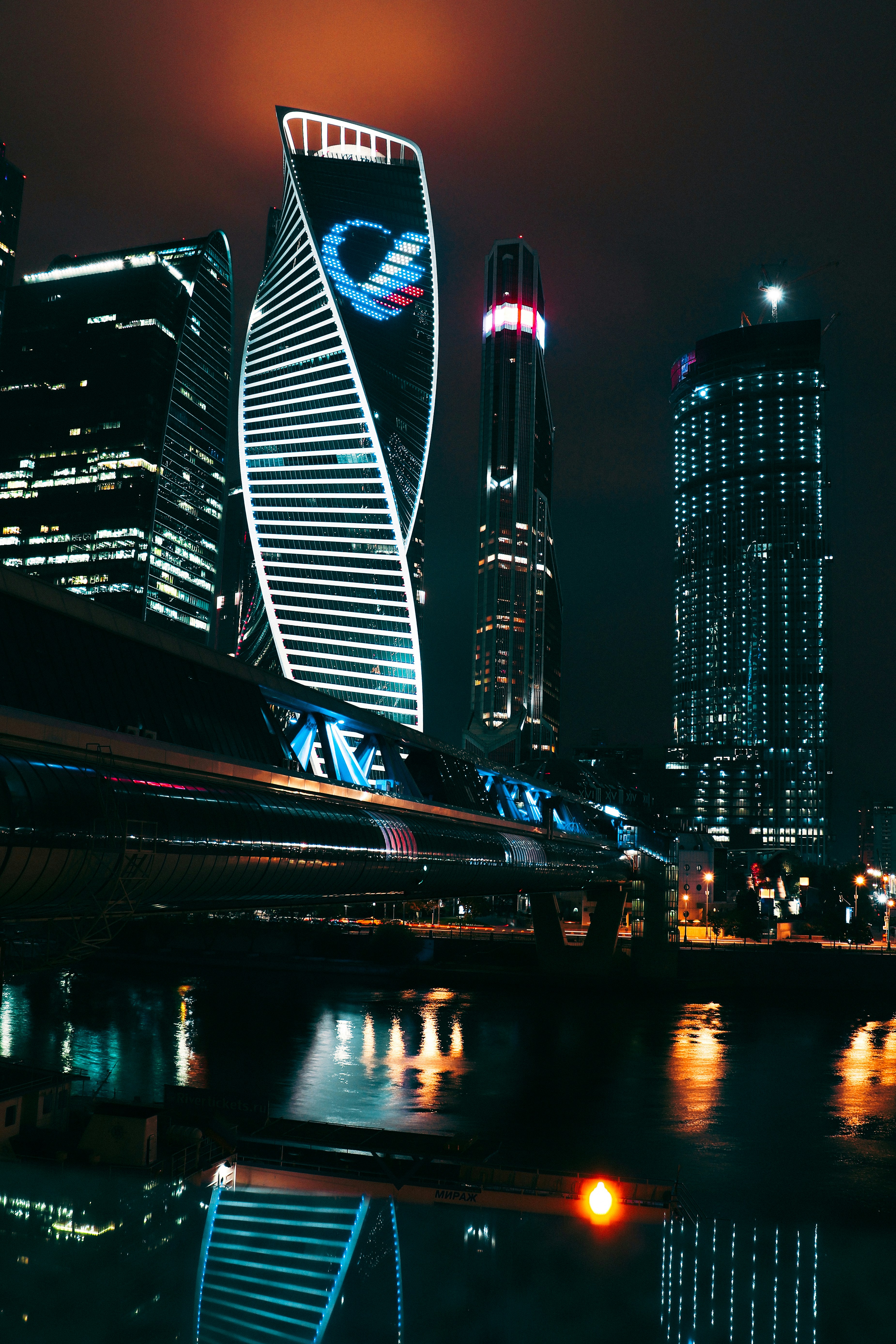 A modern skyscraper with a unique spiral design illuminated against a night sky, reflecting in the water below.