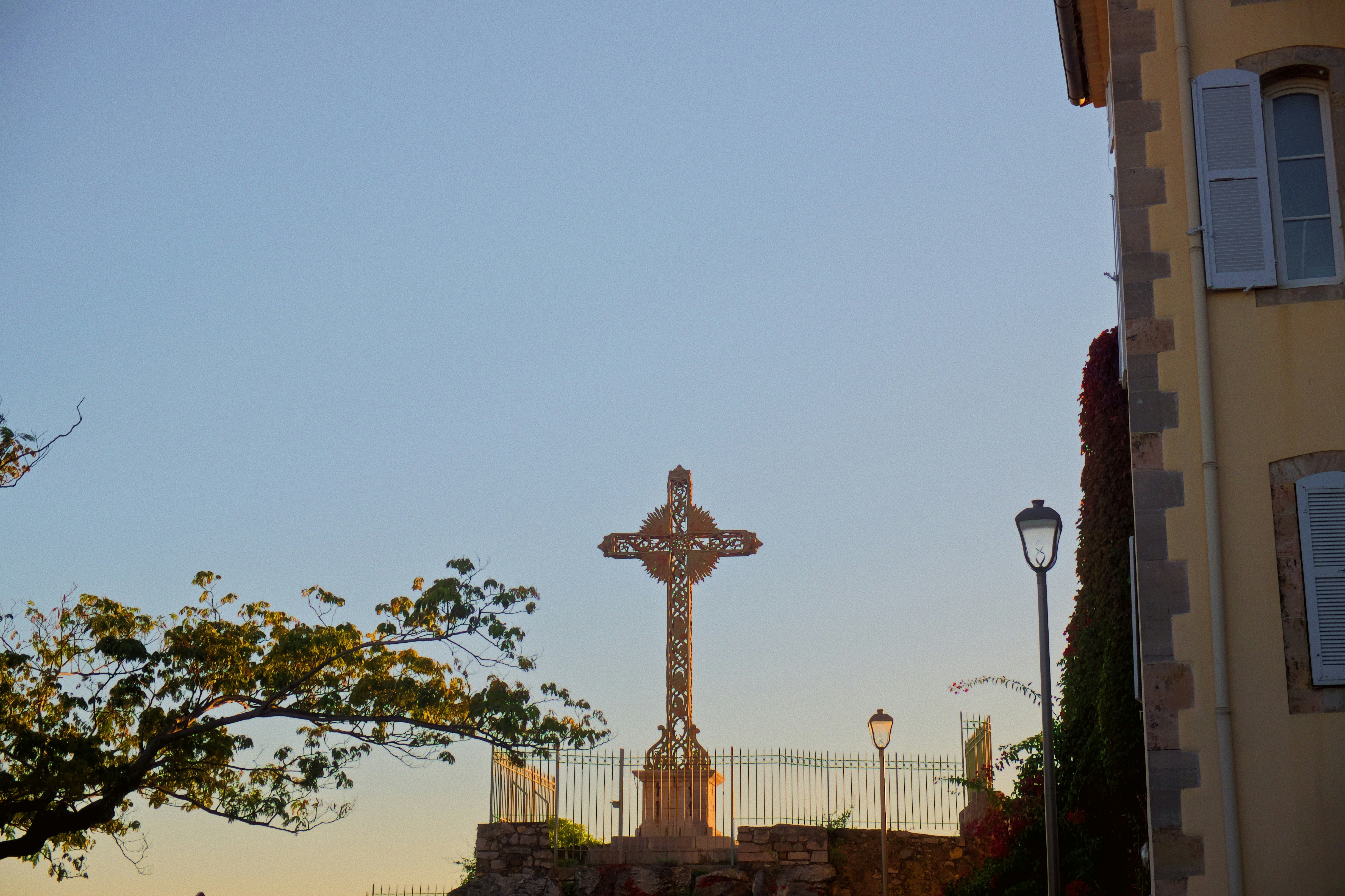 Intricate cross silhouette framed by lush foliage and a pastel sky during twilight.