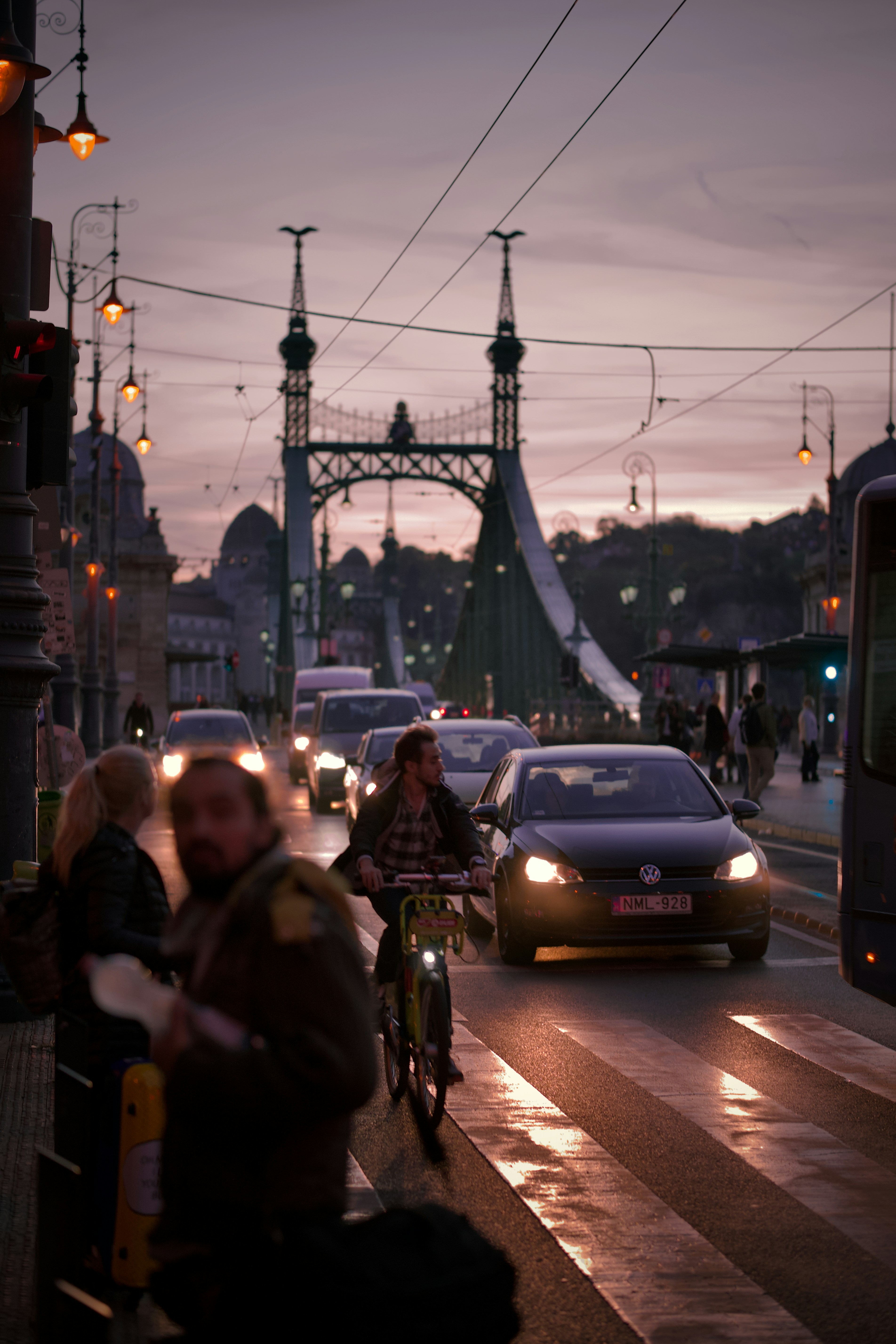 Busy street scene at dusk featuring a cyclist navigating traffic near an iconic bridge. The warm glow of streetlights contrasts with the fading light of the evening sky.