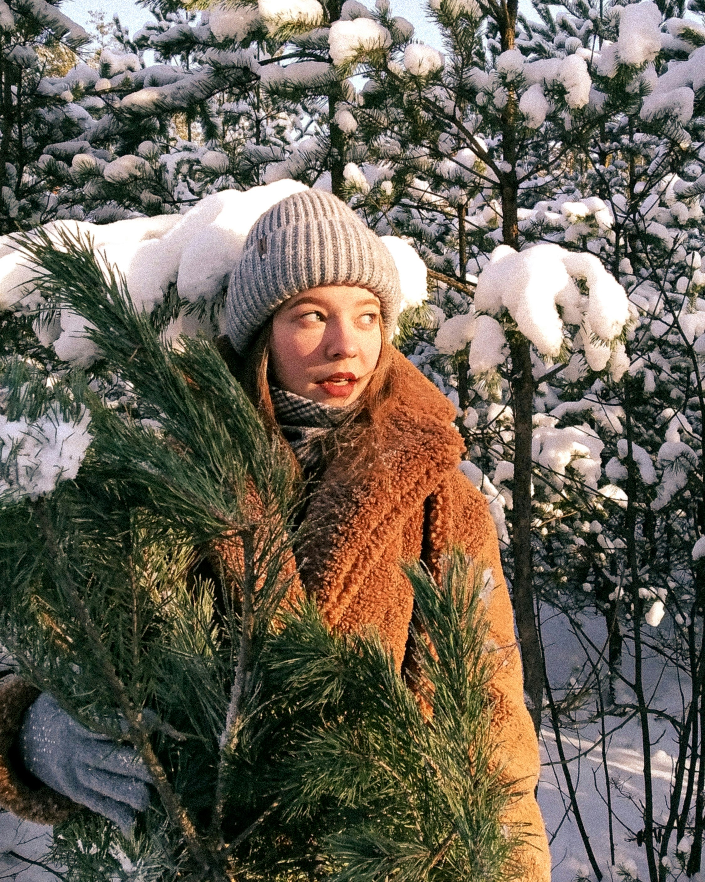 Young woman in a cozy brown coat holds evergreen branches amidst a snowy forest, with sunlight filtering through the trees.