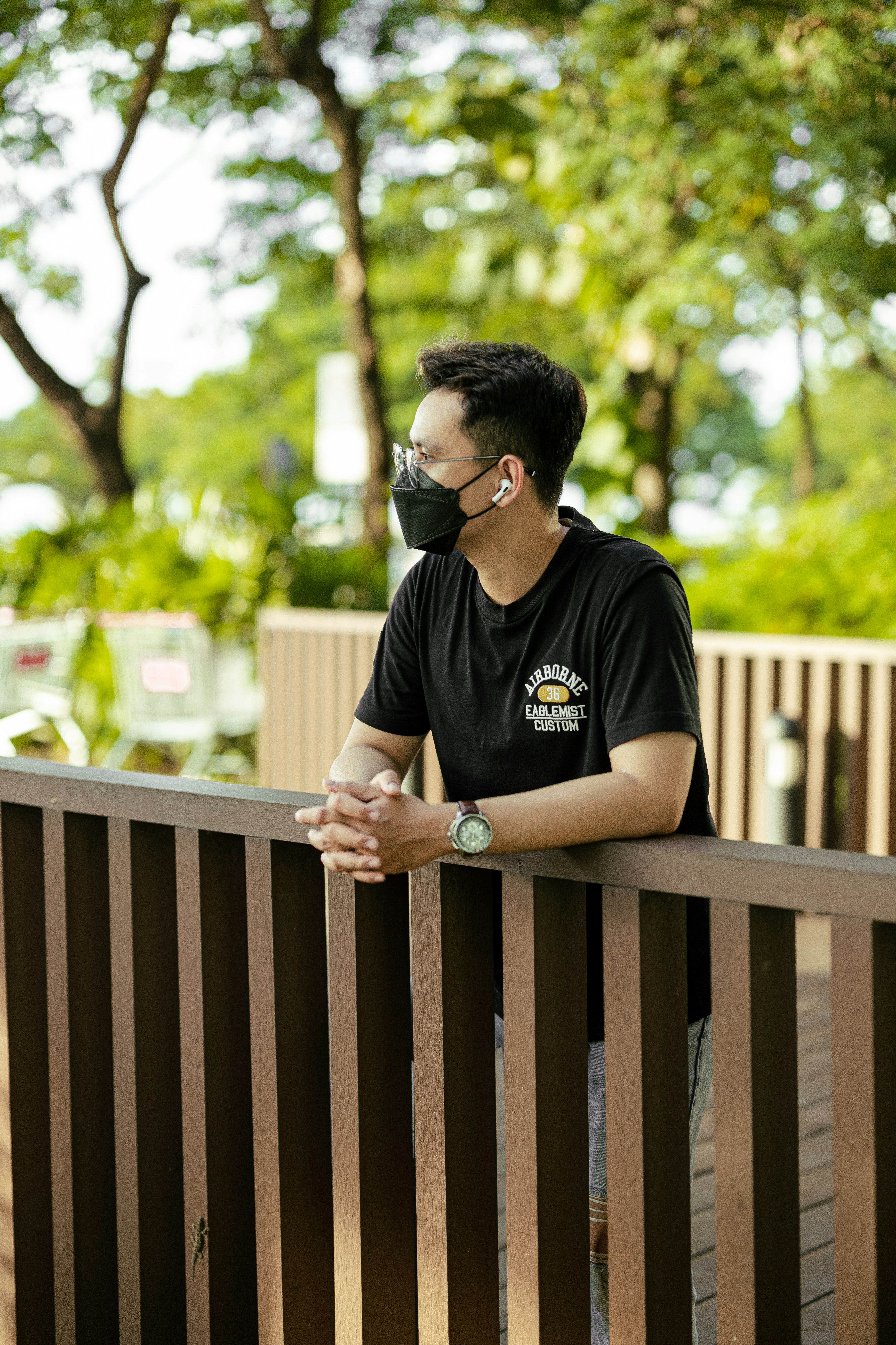 Young man in a black t-shirt and mask leans on a wooden railing, gazing thoughtfully at the lush greenery surrounding him.