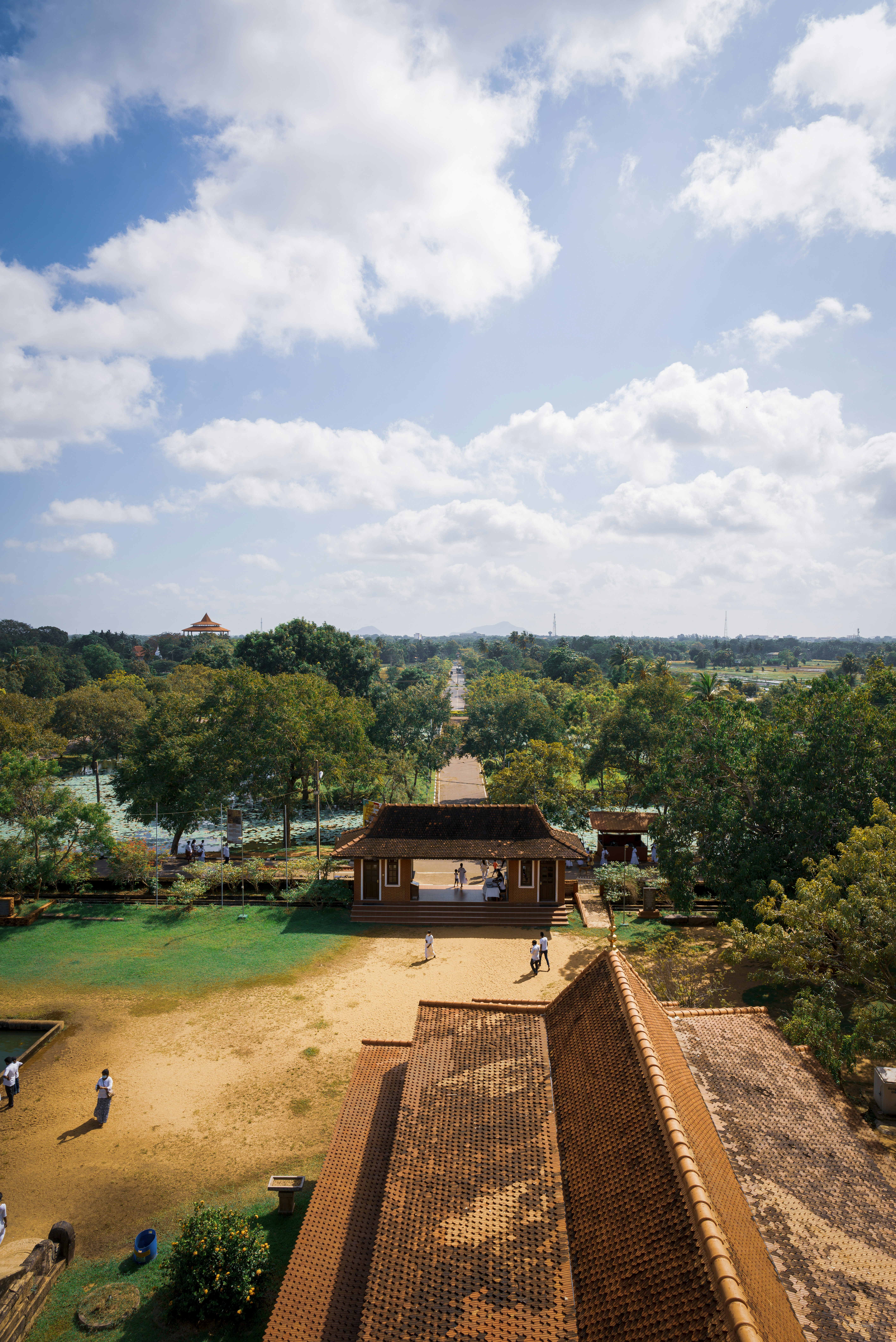 A panoramic view showcasing a blend of lush greenery and traditional architecture, with visitors exploring the serene landscape. The sky is dotted with fluffy clouds, enhancing the tranquil atmosphere.
