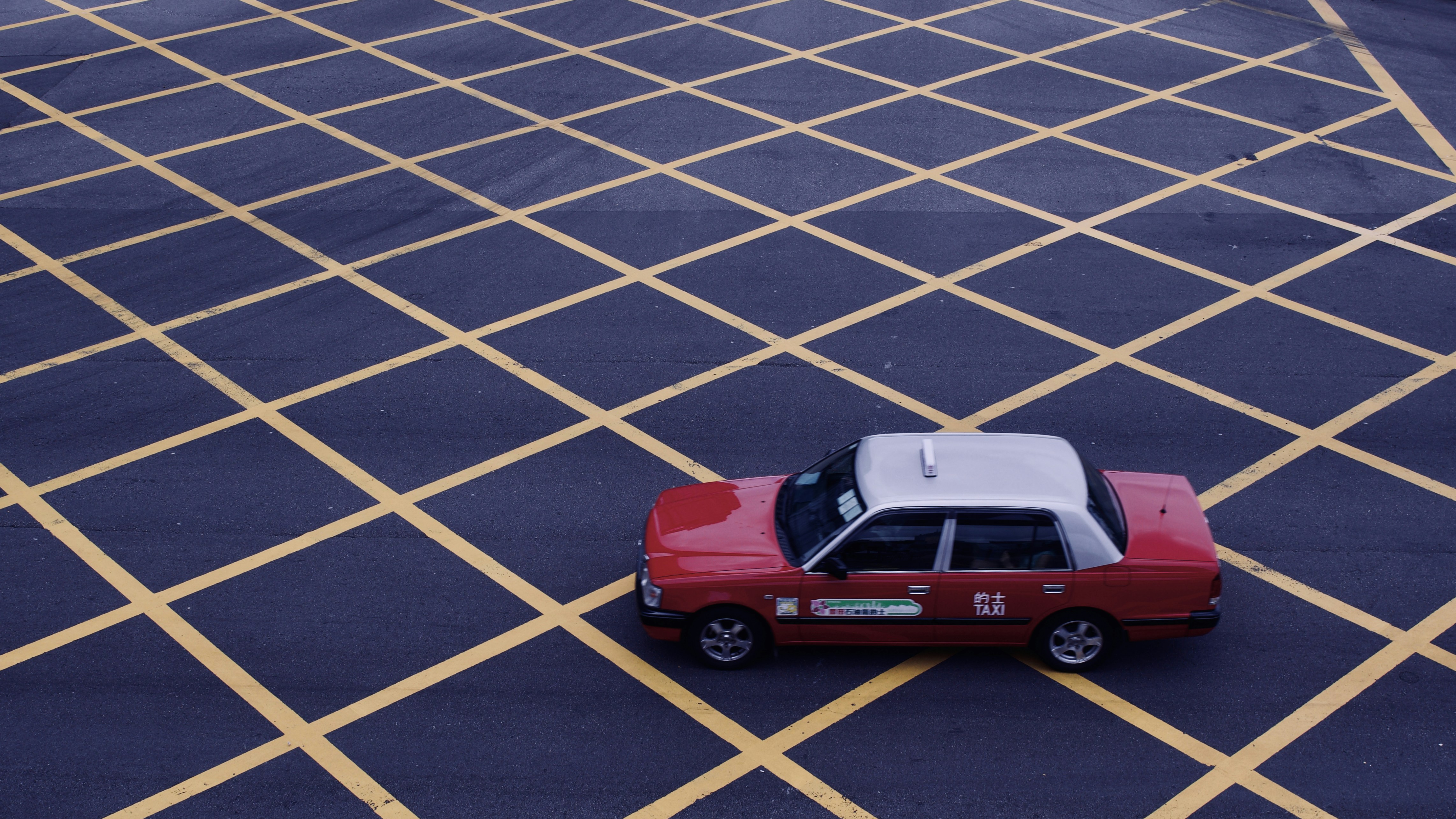 A red taxi navigates a geometric pattern of yellow lines on an asphalt surface, highlighting the interplay between urban transportation and city design.