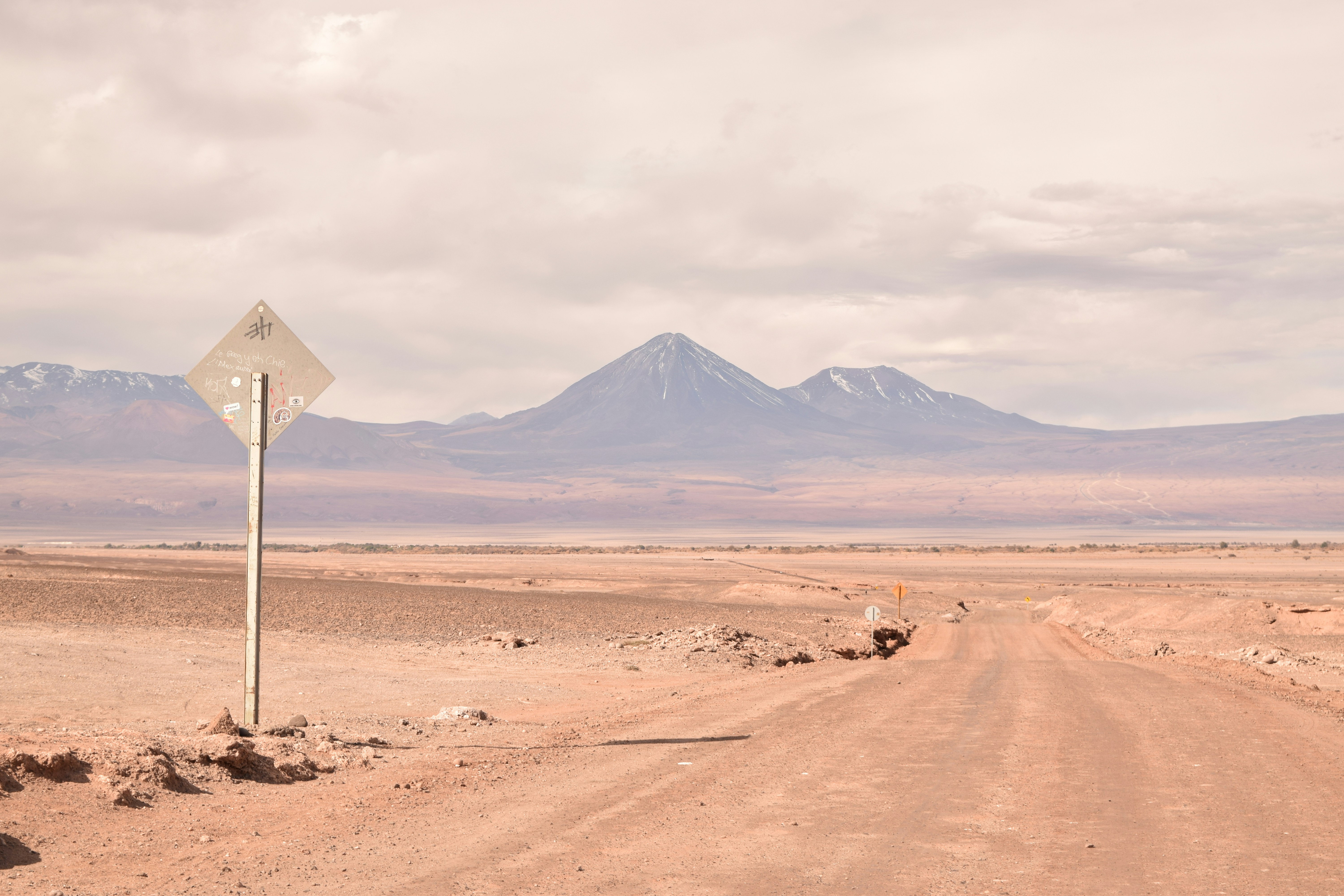A rugged dirt road leads towards distant snow-capped mountains under a cloudy sky, with a weathered road sign marking the way.