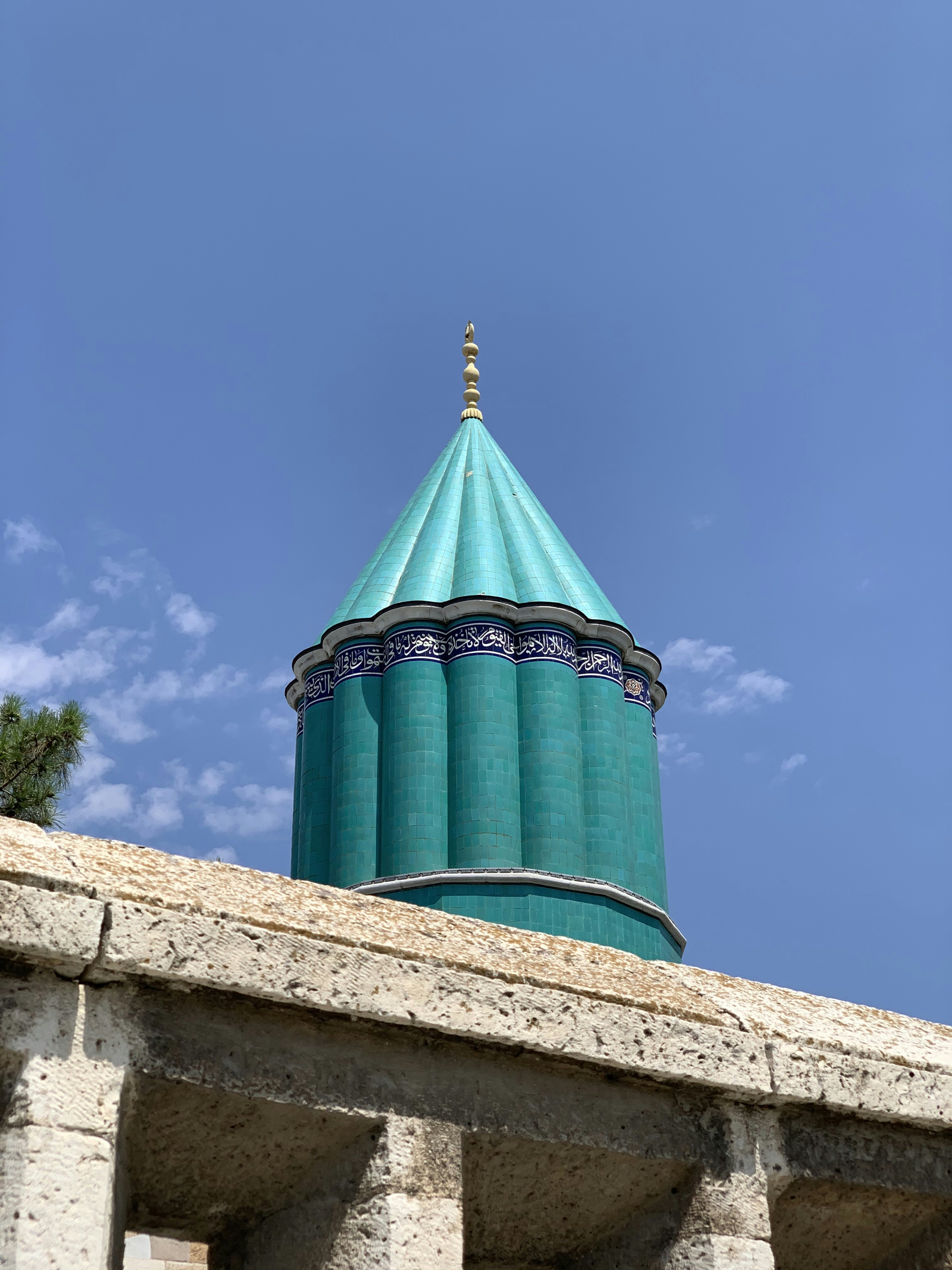 The striking turquoise dome of a historical structure rises against a clear blue sky, framed by a textured stone wall in the foreground.